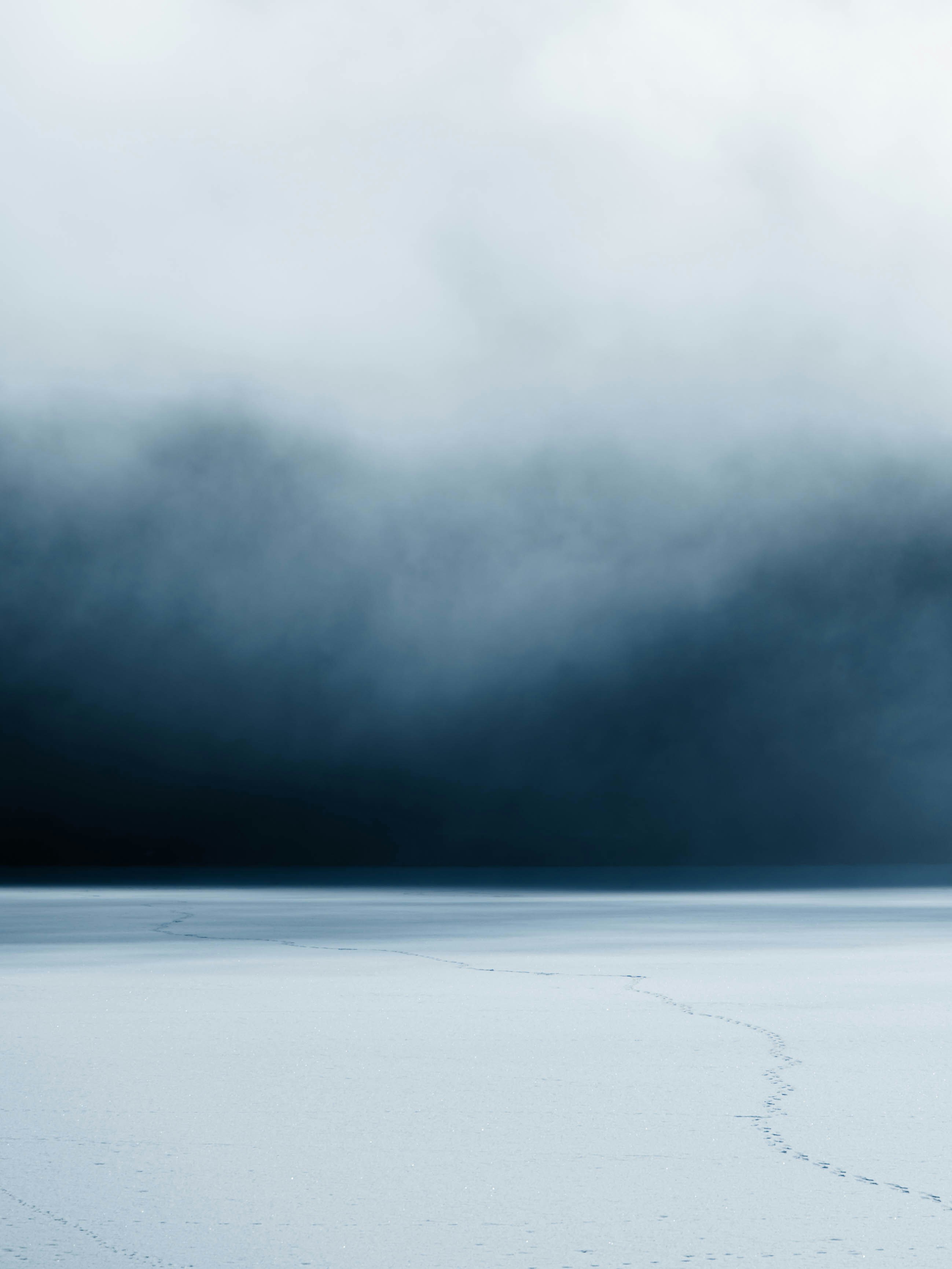 Minimalist winter landscape with footprints on snow leading into the distance, under dramatic foggy sky | Misty, cold landscape with footprints in snow