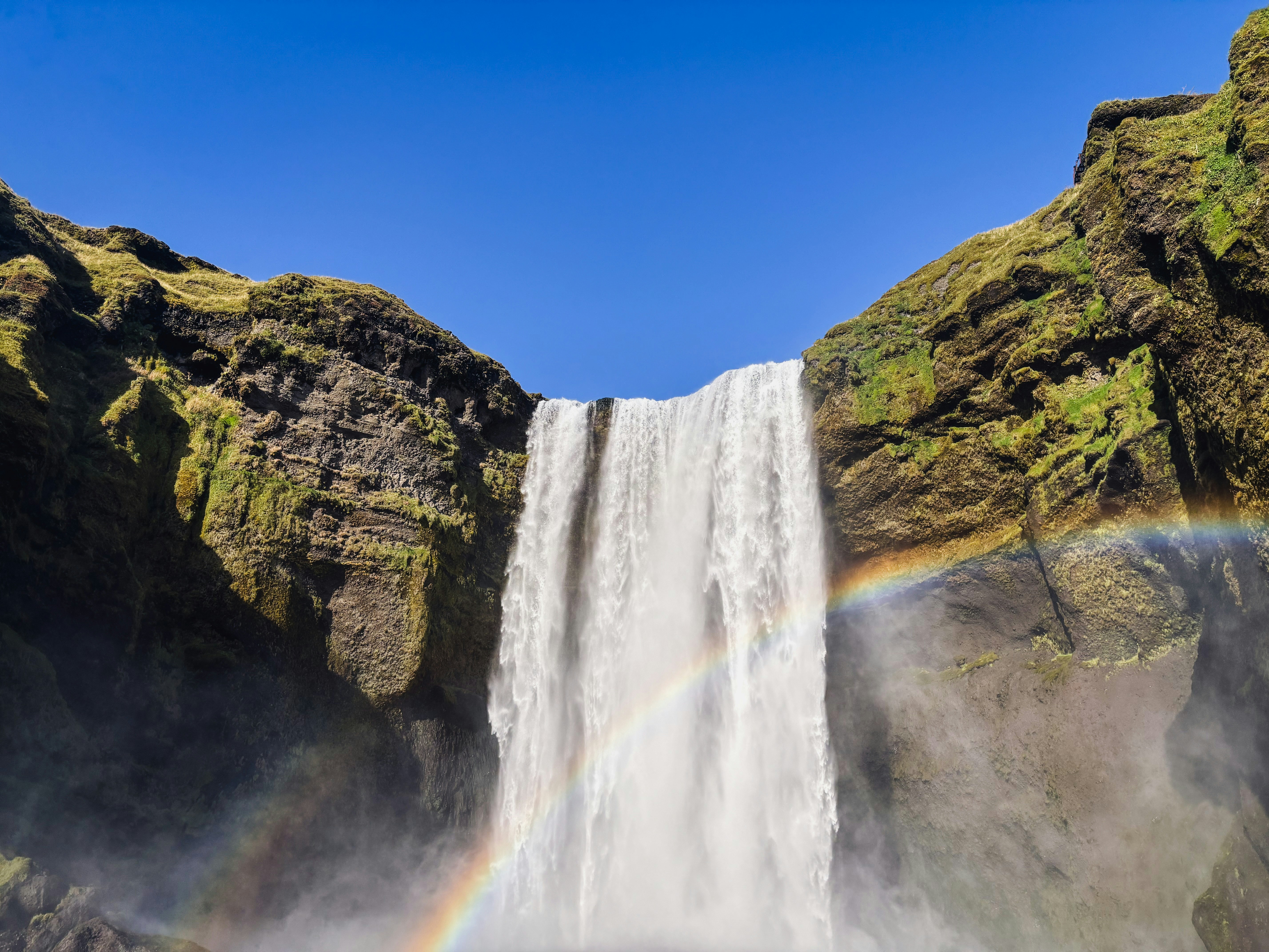 Majestic waterfall cascading down rocky cliffs, with a vibrant rainbow arching across the mist. The scene captures the beauty of nature's interplay between water and sunlight.
