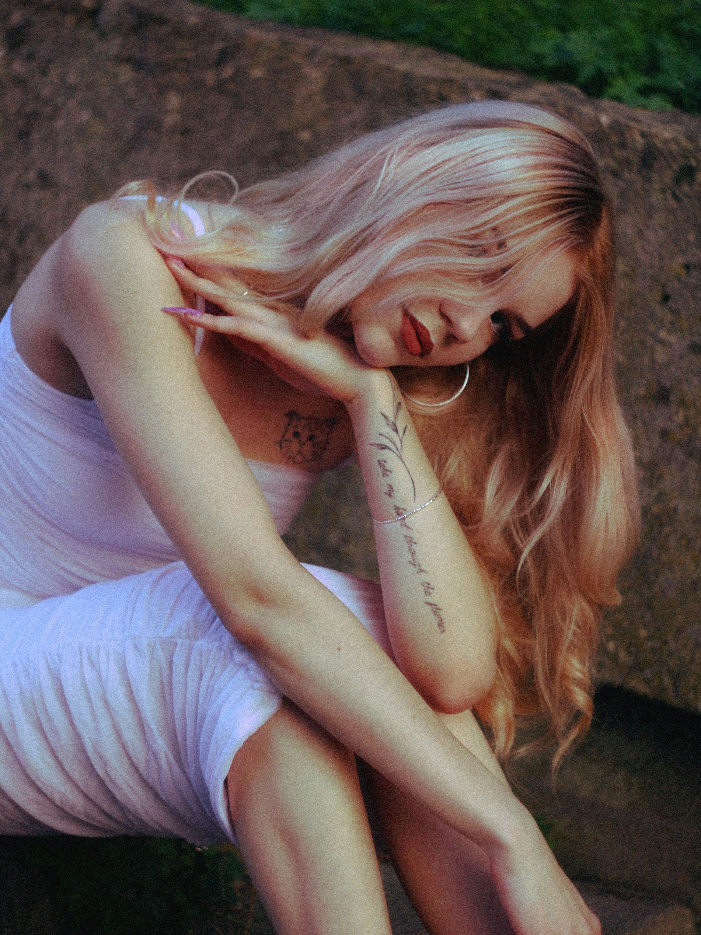 A contemplative young woman with flowing blonde hair rests her chin on her hand, adorned with delicate tattoos, against a textured backdrop of stone and greenery.