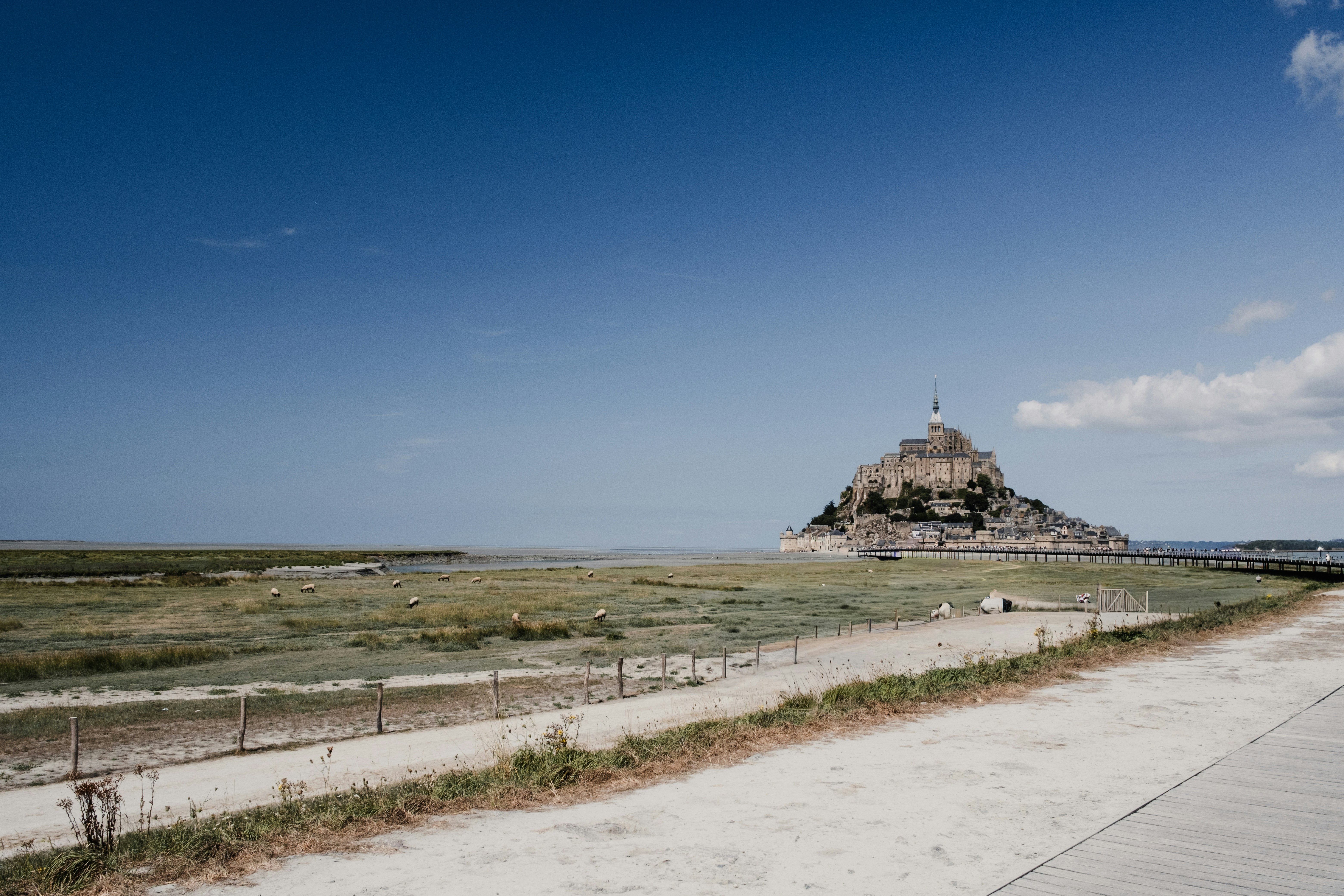 Mont Saint-Michel rises majestically from the surrounding landscape, showcasing its medieval architecture against a clear blue sky. The serene path invites exploration.