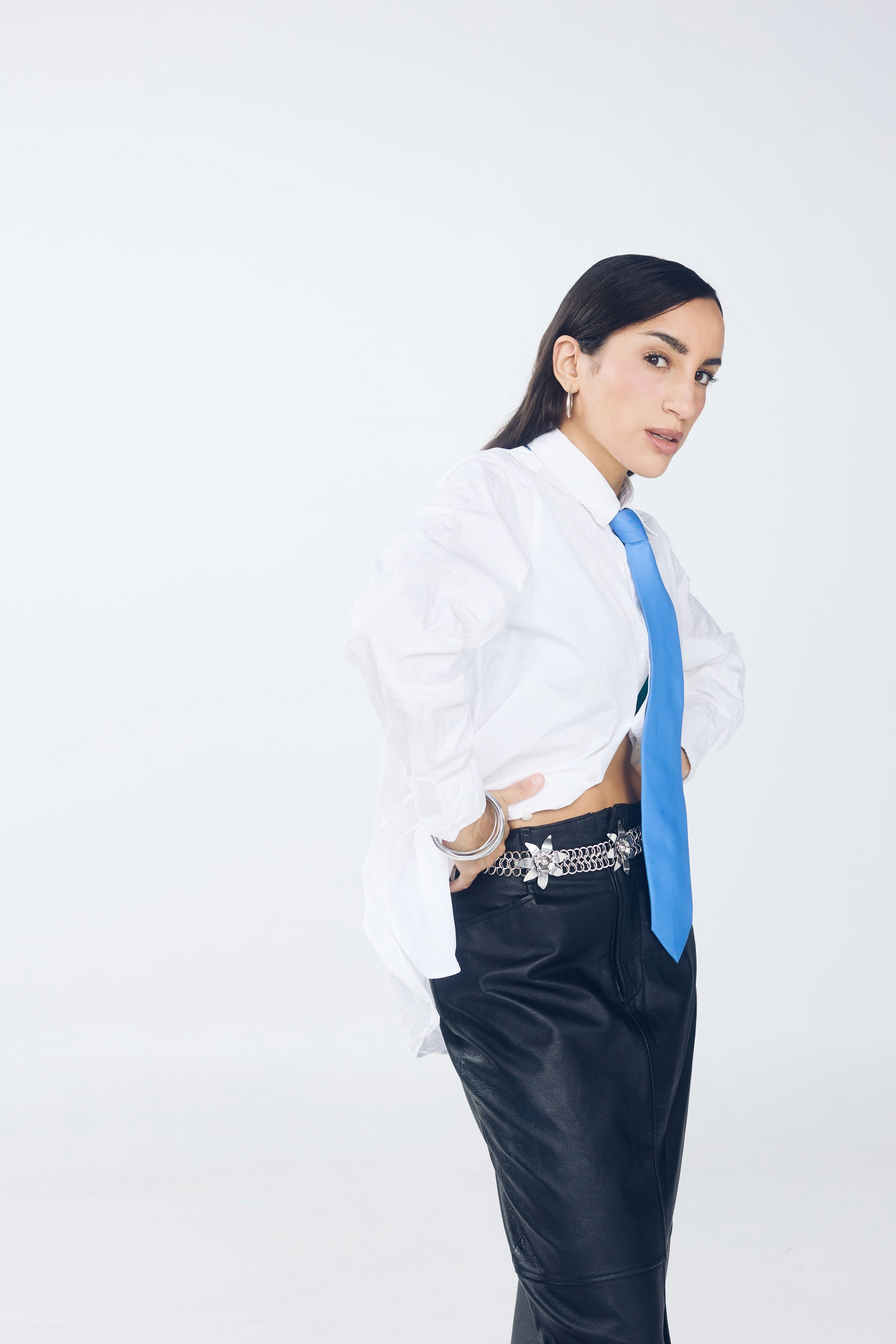 A confident individual poses in a stylish outfit featuring a white shirt and a blue tie, set against a minimalist background.