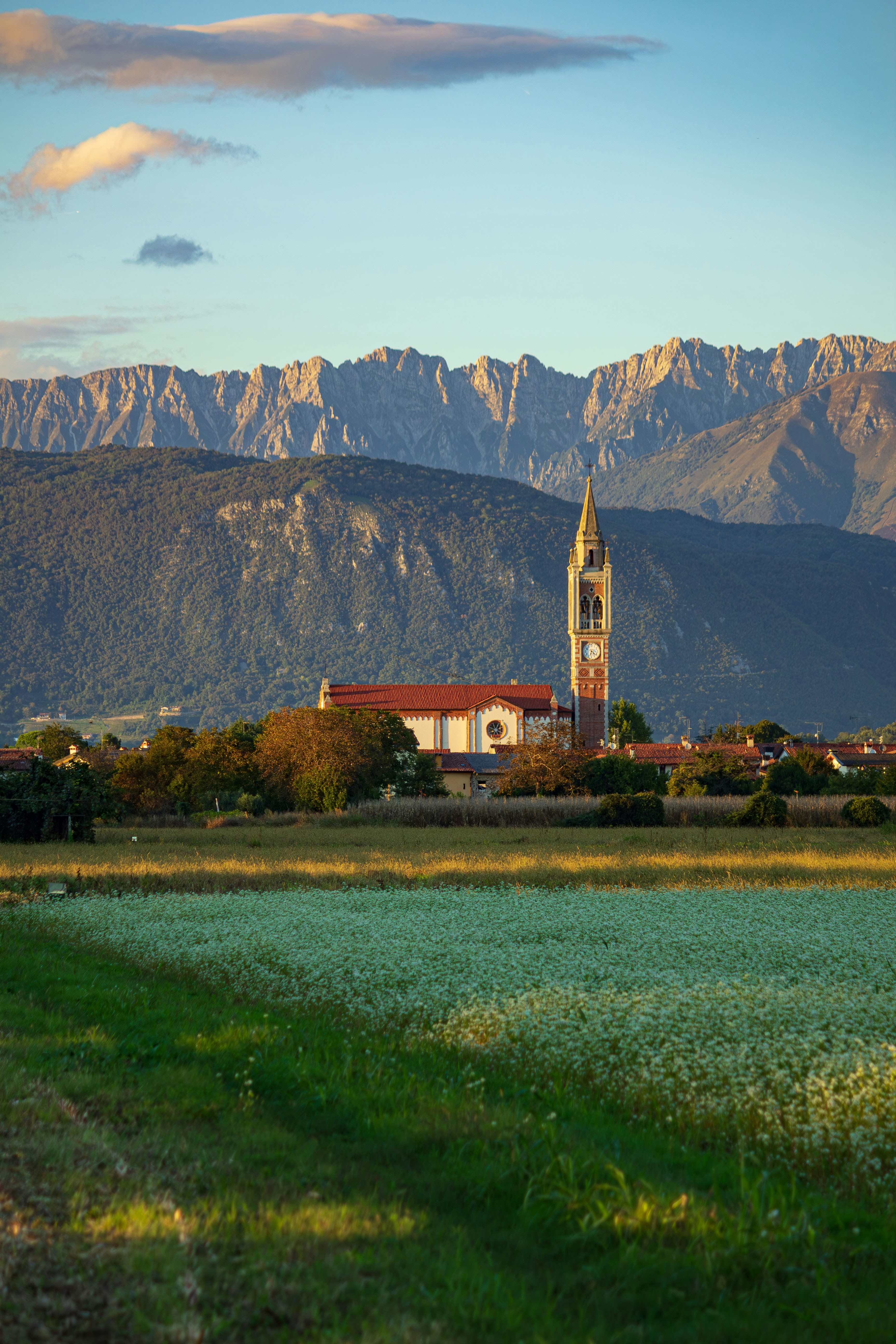Chiesa dei Santi Ilario e Tiziano (Reana del Rojale, IT - 2025) | Village church with bell tower against mountain backdrop.