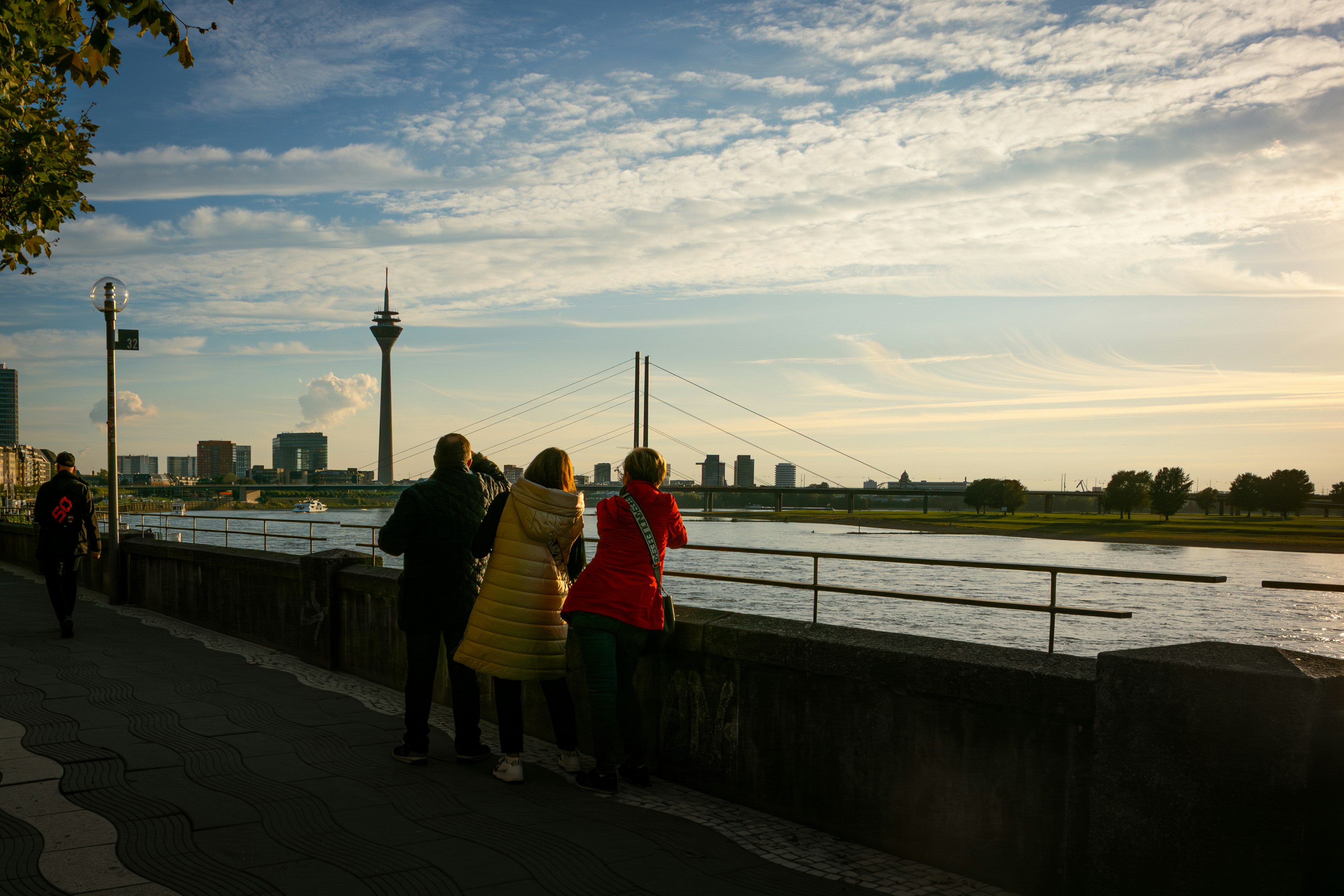 Three individuals admire the river view while standing on a promenade, with a city skyline and a tower in the background. The scene is illuminated by the warm glow of the setting sun.