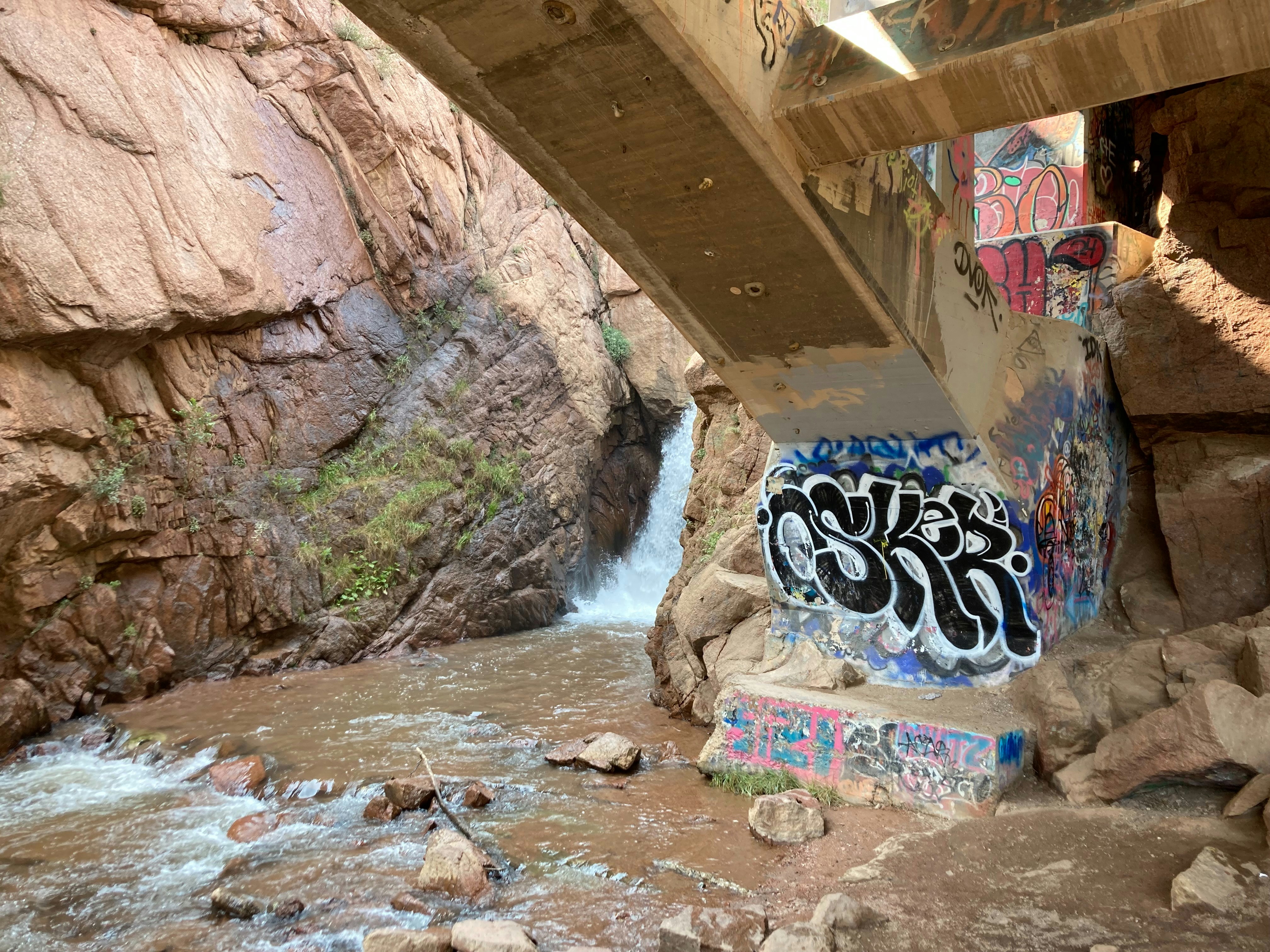 Rainbow Falls also known as Graffiti Falls. The graffiti on the walls along the waterfalls were removed. There are still graffitis left on the bridge over the waterfalls. | Waterfall under a graffiti-covered bridge in a canyon