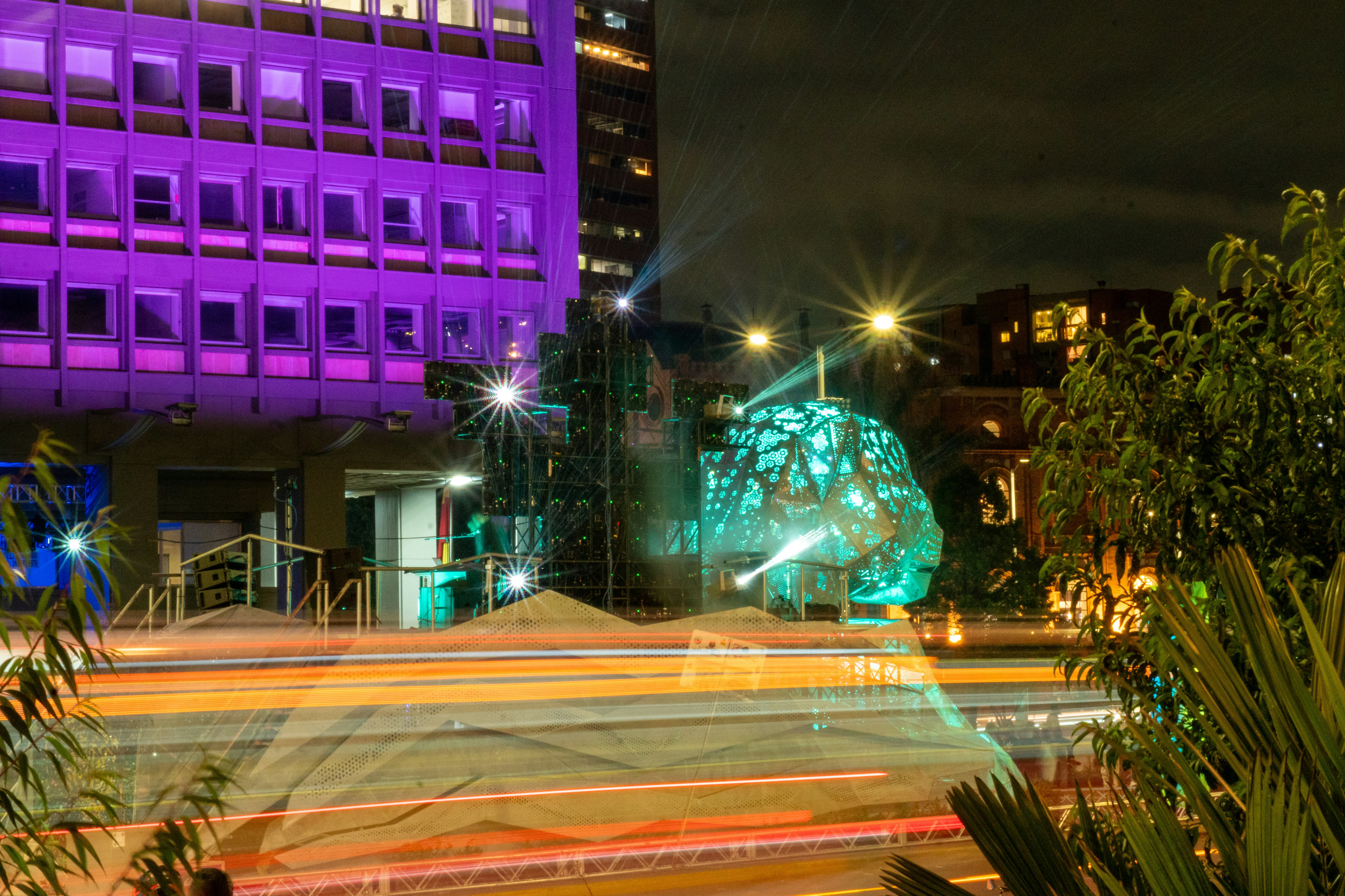 A vibrant sculpture glows in teal amidst a backdrop of a modern building illuminated in purple, capturing the essence of urban nightlife.