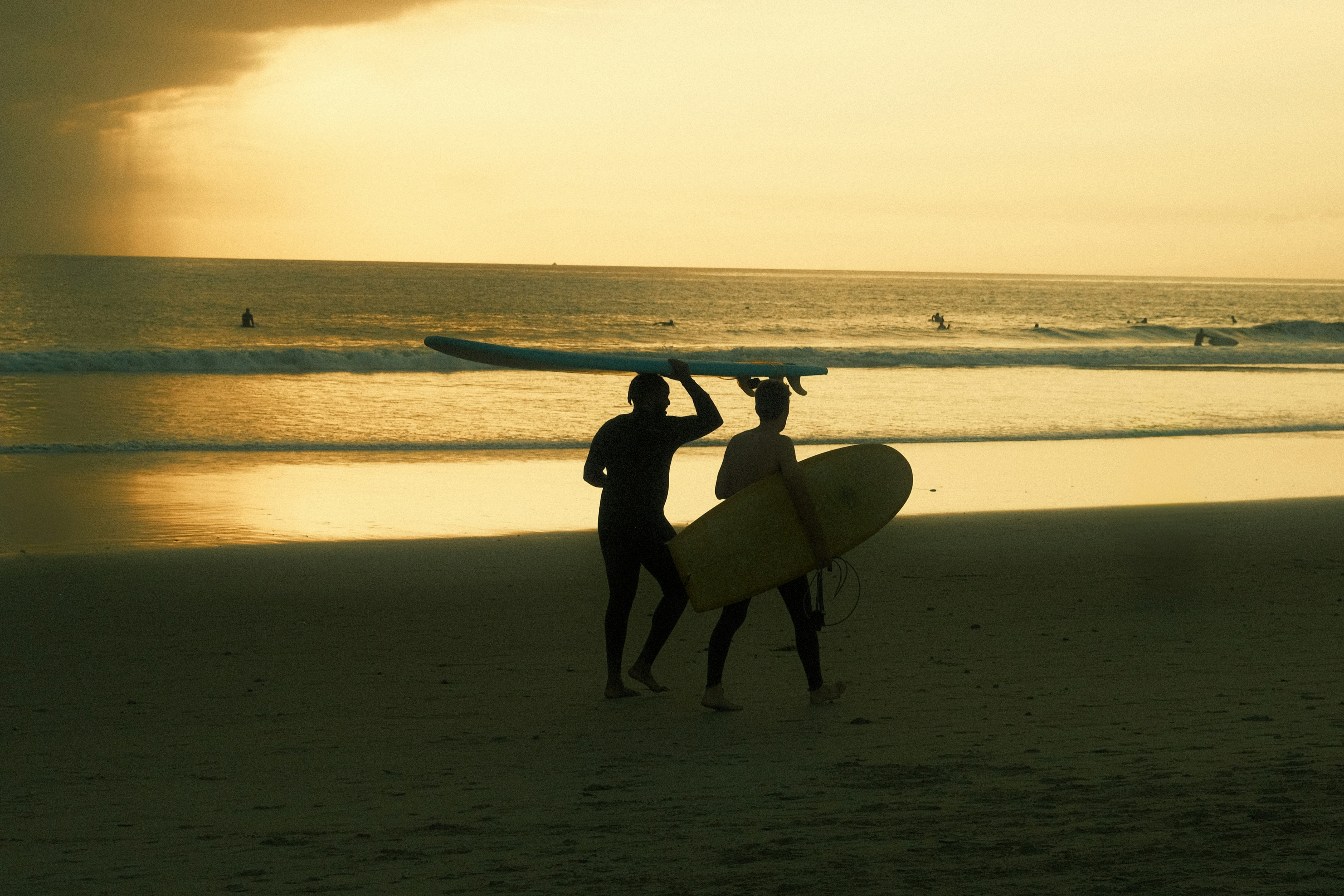 Two surfers walk on the beach at sunset.