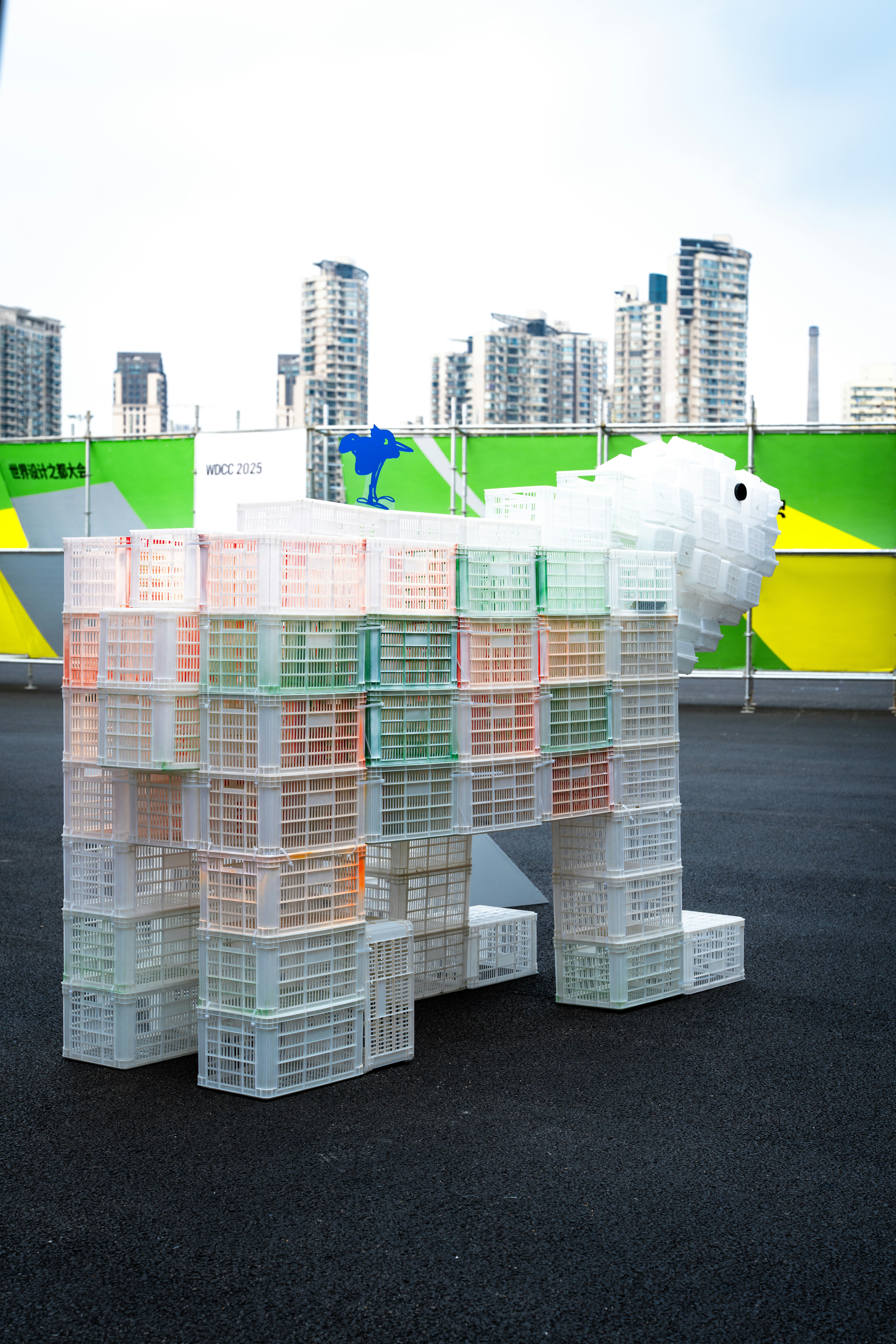 A large dog sculpture made from colorful plastic crates stands in an urban setting, showcasing creativity and sustainability. The backdrop features modern buildings and vibrant signage.