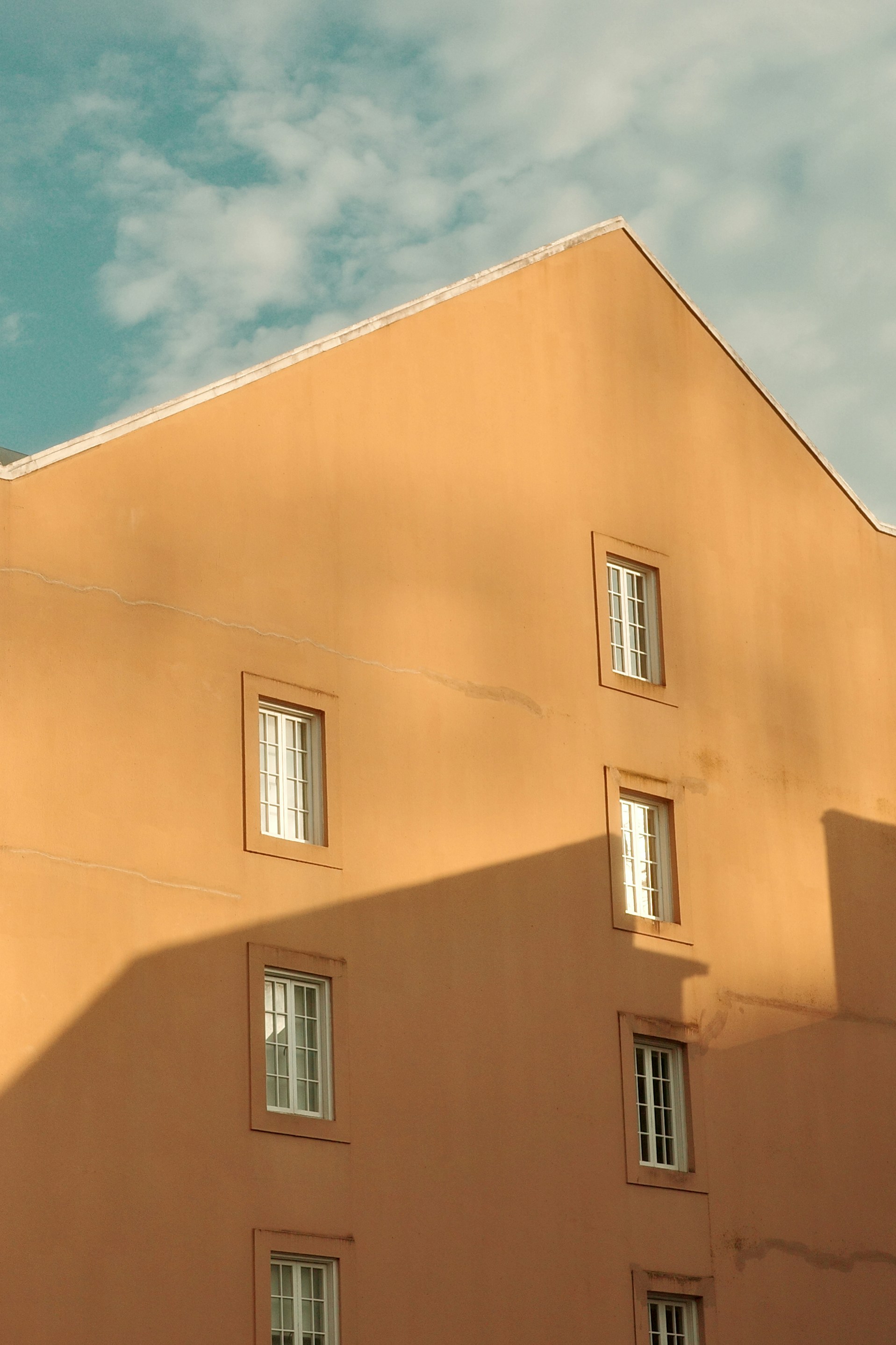 Shadows and sunshine on a bright yellow-orange building with clouds in the background | Warm building facade with windows and sky