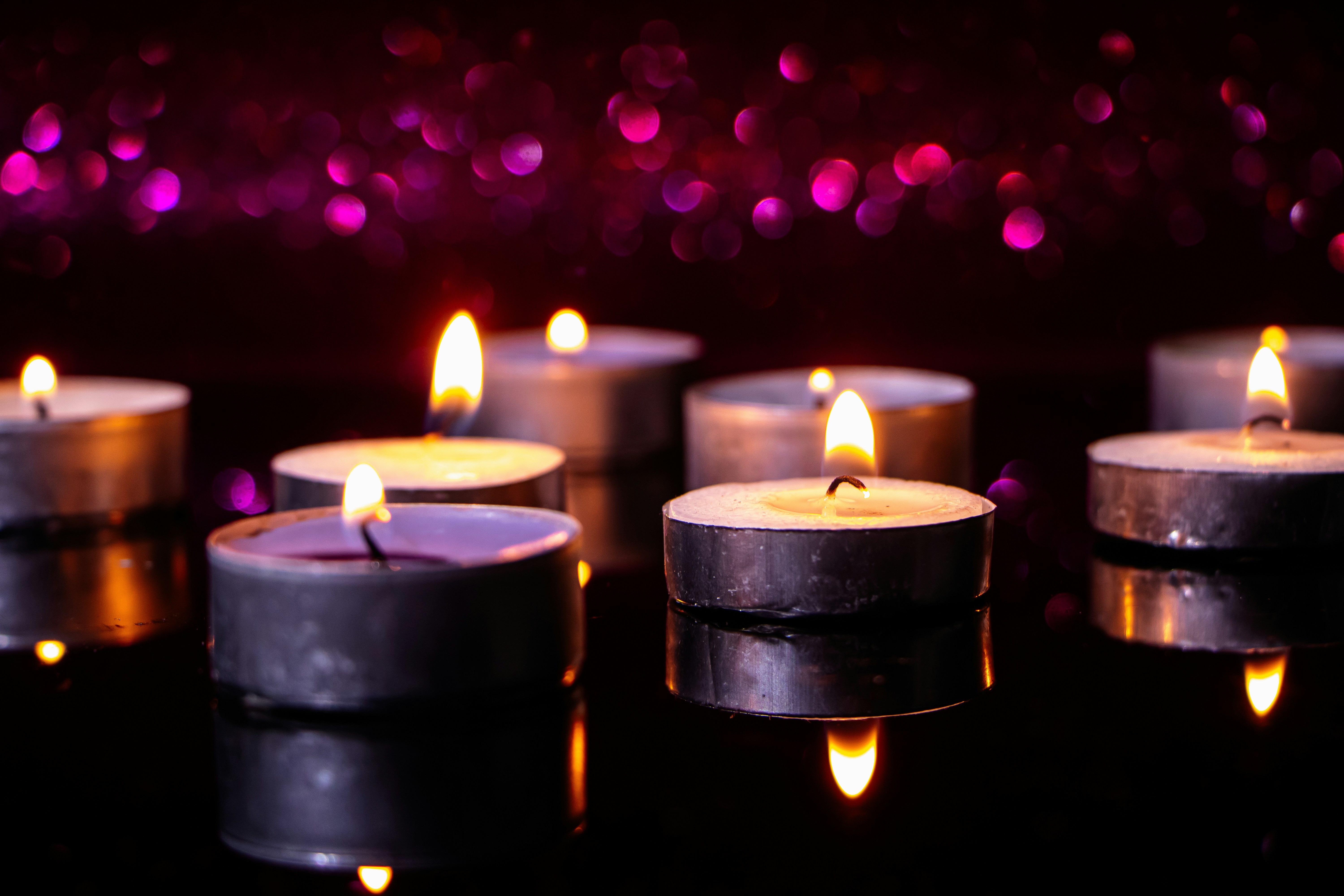 A serene arrangement of glowing tealight candles on a reflective surface, set against a dreamy backdrop of purple and pink bokeh lights. A warm, meditative ambiance that evokes calm, romance, and quiet beauty. | Several lit candles on a reflective surface with bokeh lights.