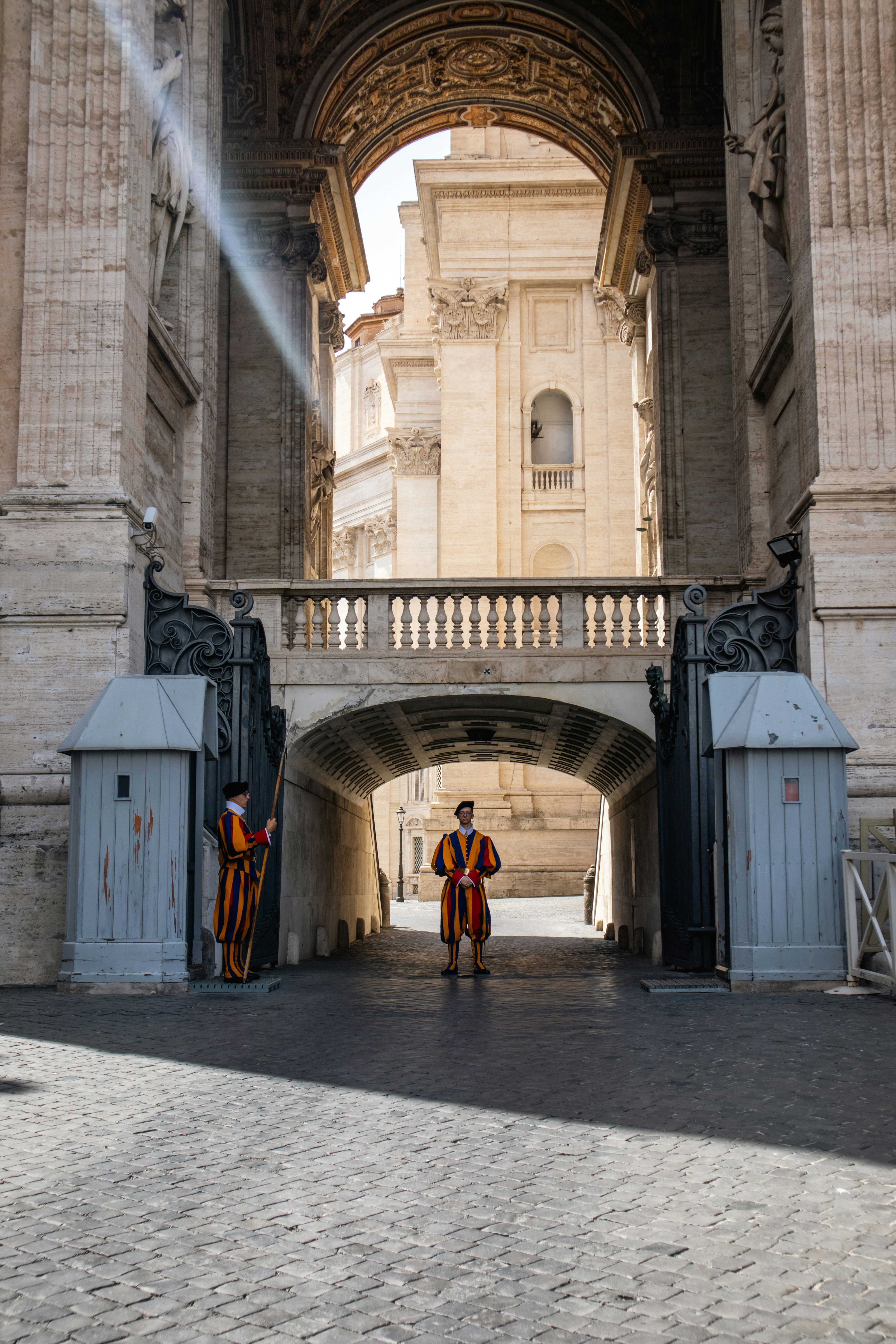 Two Swiss Guards in traditional uniforms stand at the entrance of a grand archway leading to the Vatican, showcasing the architectural beauty and historical significance of the site.