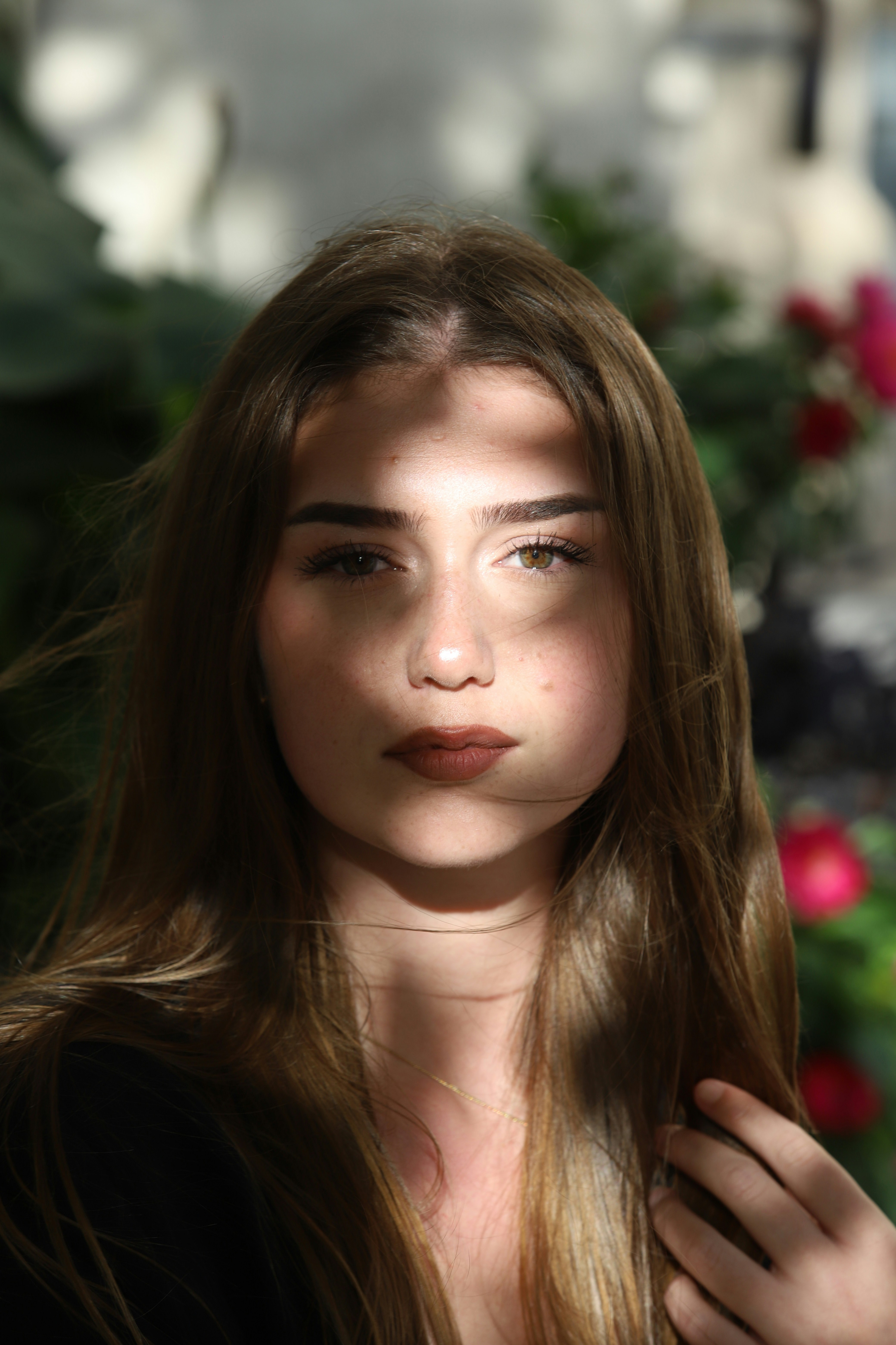 A young woman stands amidst vibrant foliage, her face partially illuminated by soft shadows, creating a striking contrast with her natural beauty.