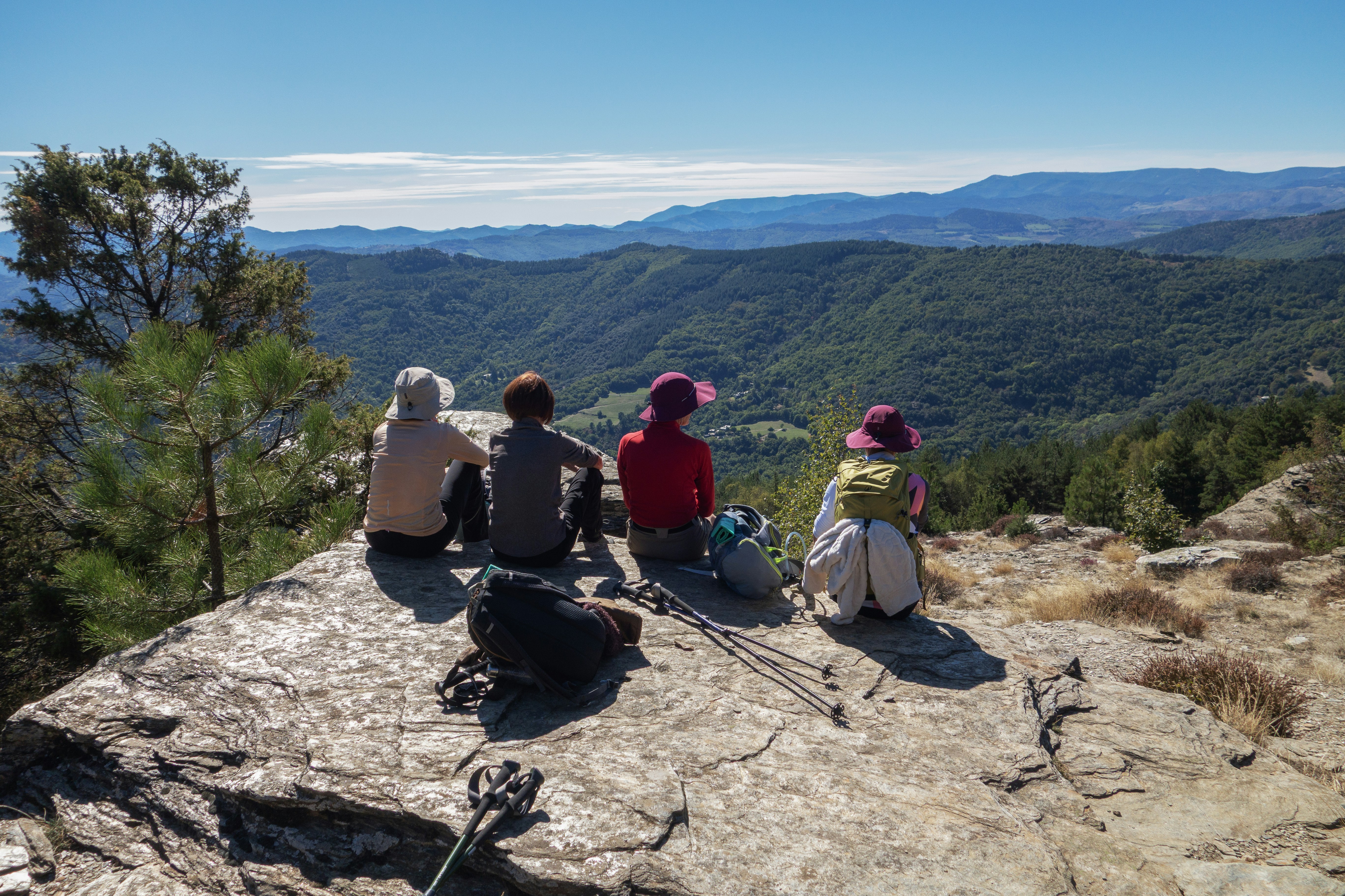Four hikers pause on a rocky outcrop, gazing at the expansive landscape of rolling hills and distant mountains. The scene captures a tranquil moment in nature.