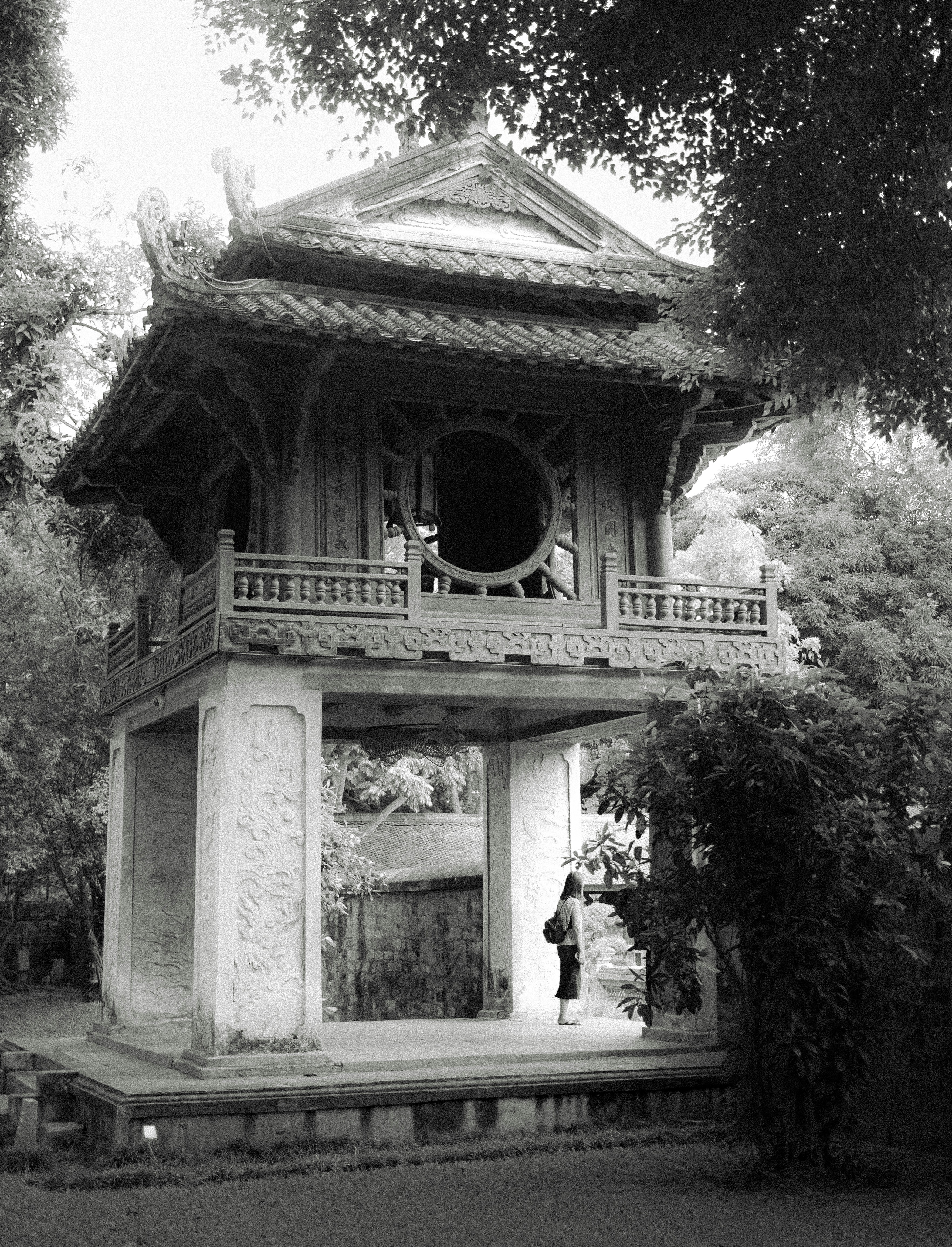 A traditional pagoda-style structure surrounded by lush greenery, showcasing intricate wooden carvings and a circular opening. A solitary figure stands nearby, enhancing the sense of scale.
