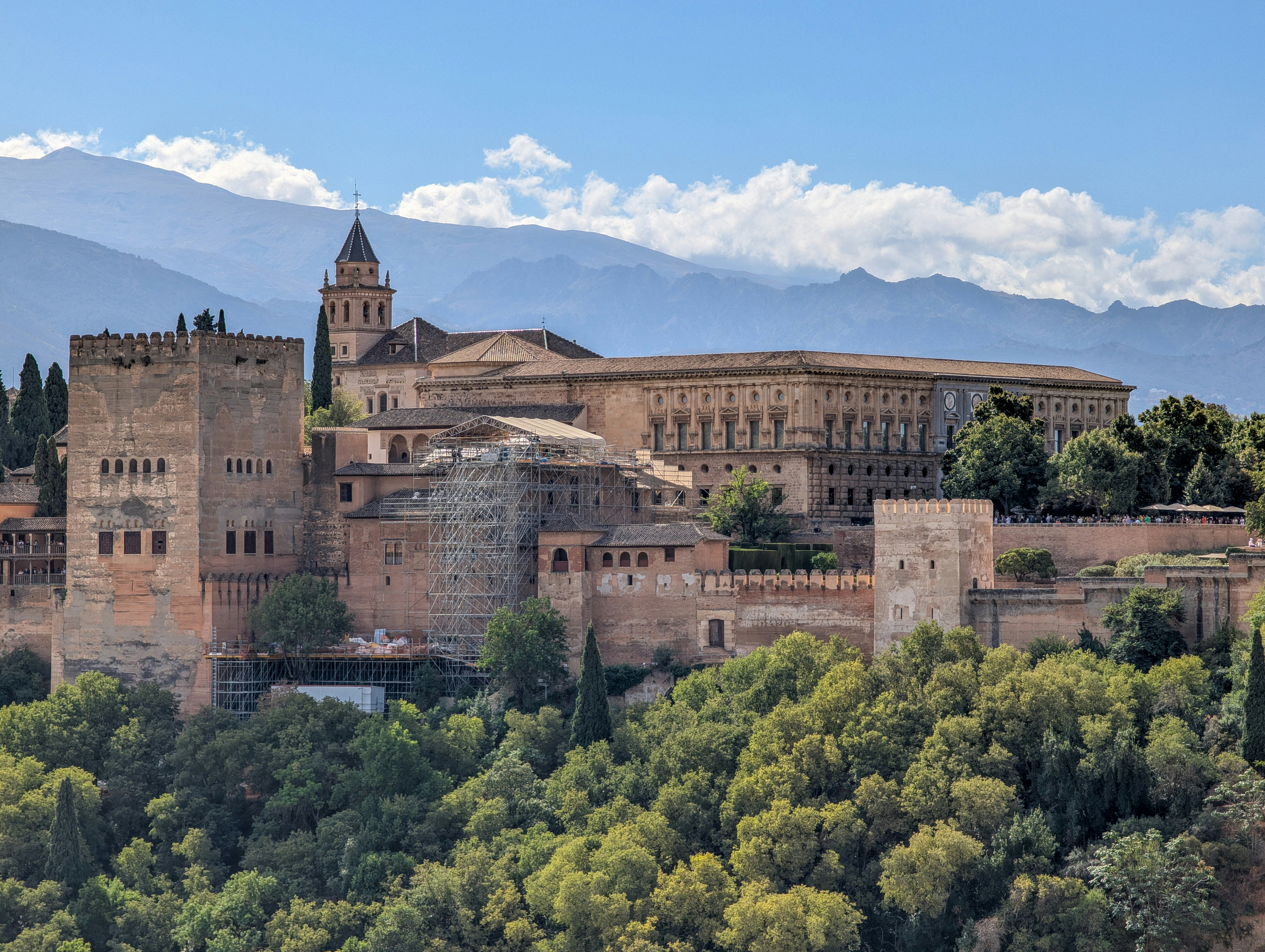 The Alhambra fortress complex rises gracefully amidst lush greenery with distant mountains framing the scene. A blend of architectural grandeur and natural beauty.