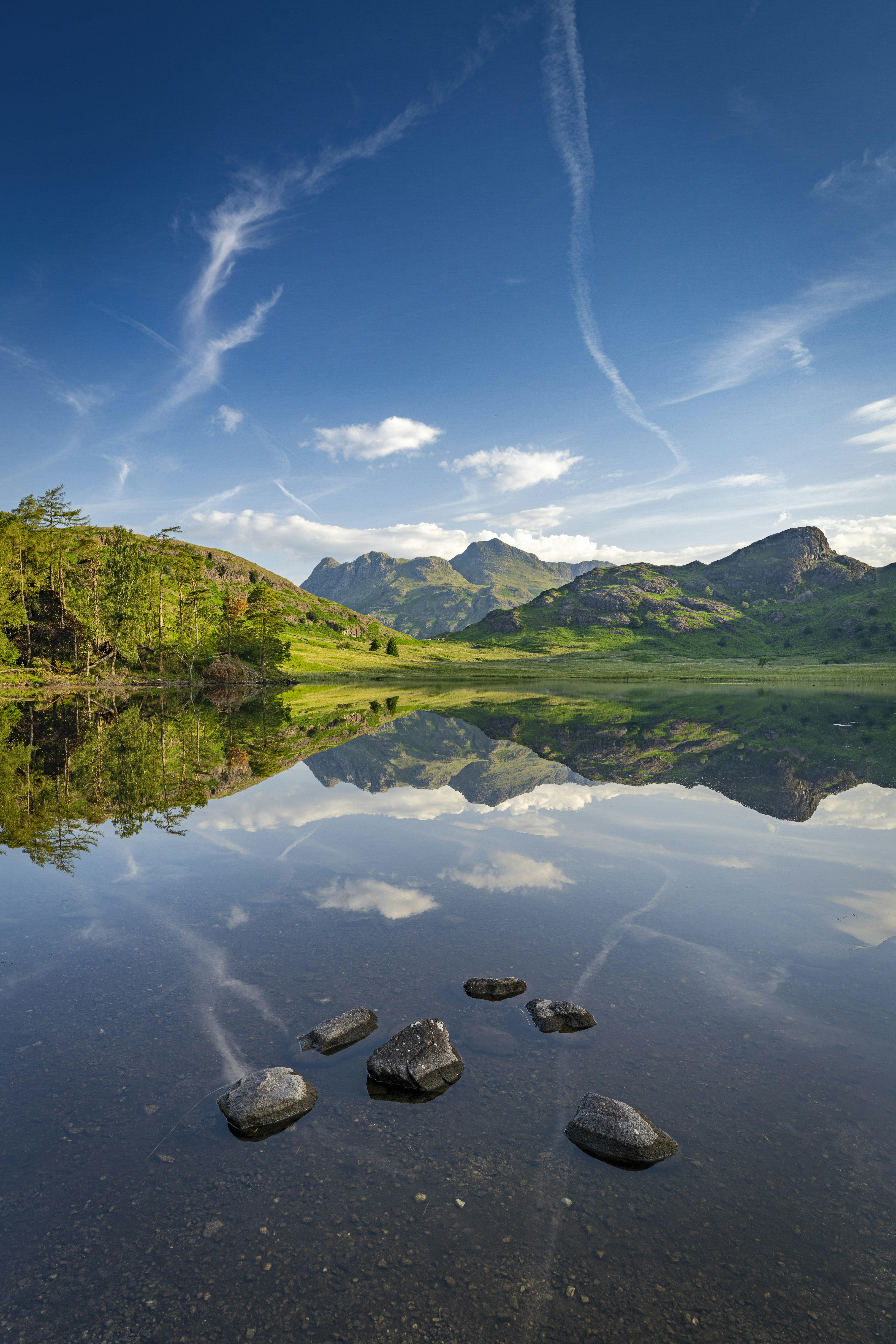 Calm lake reflecting lush green hills and a blue sky with wispy clouds, accented by scattered stones in the foreground.