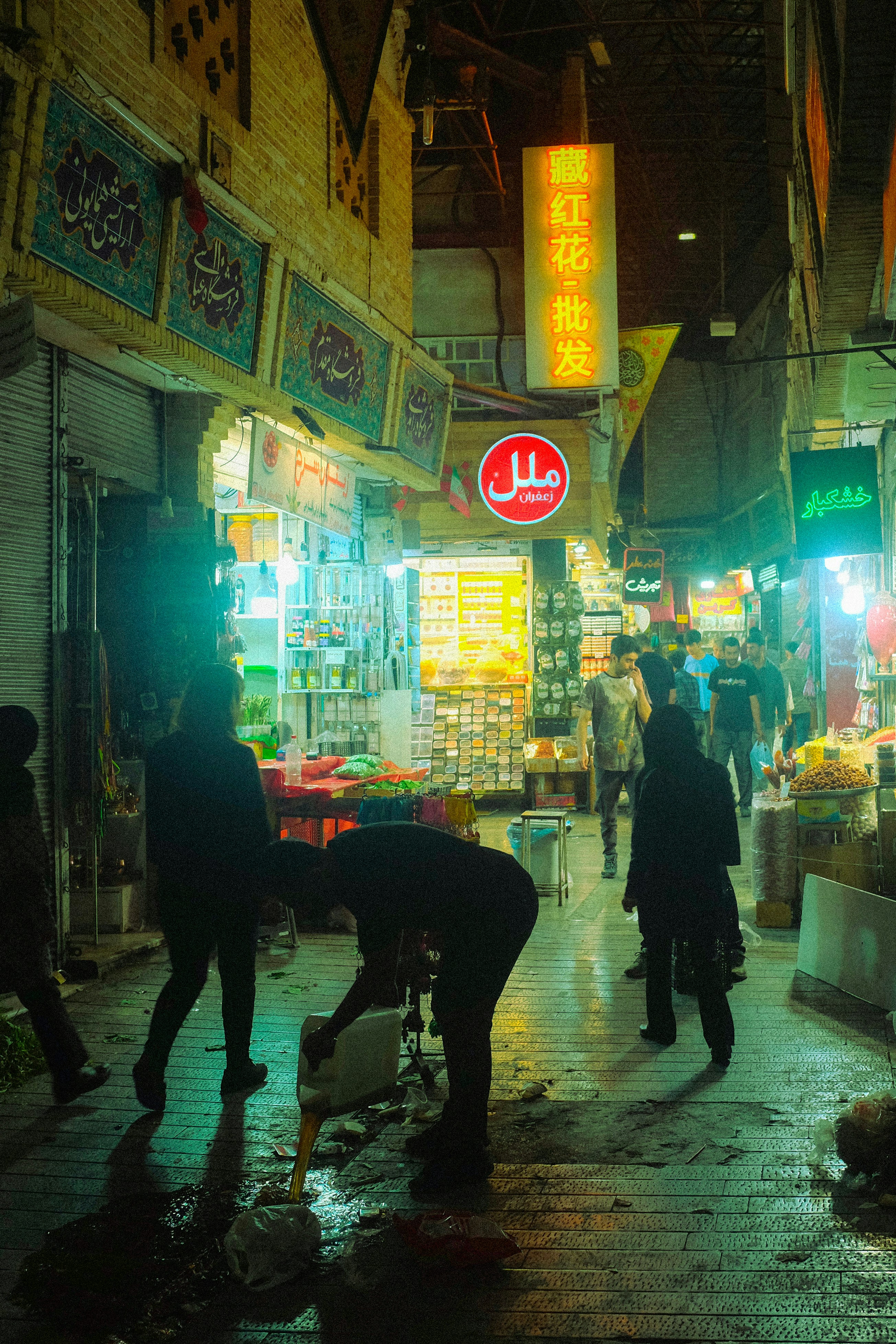 Vibrant night market scene with illuminated shops and figures engaging in daily activities. A vendor pours liquid onto the ground amidst colorful displays.