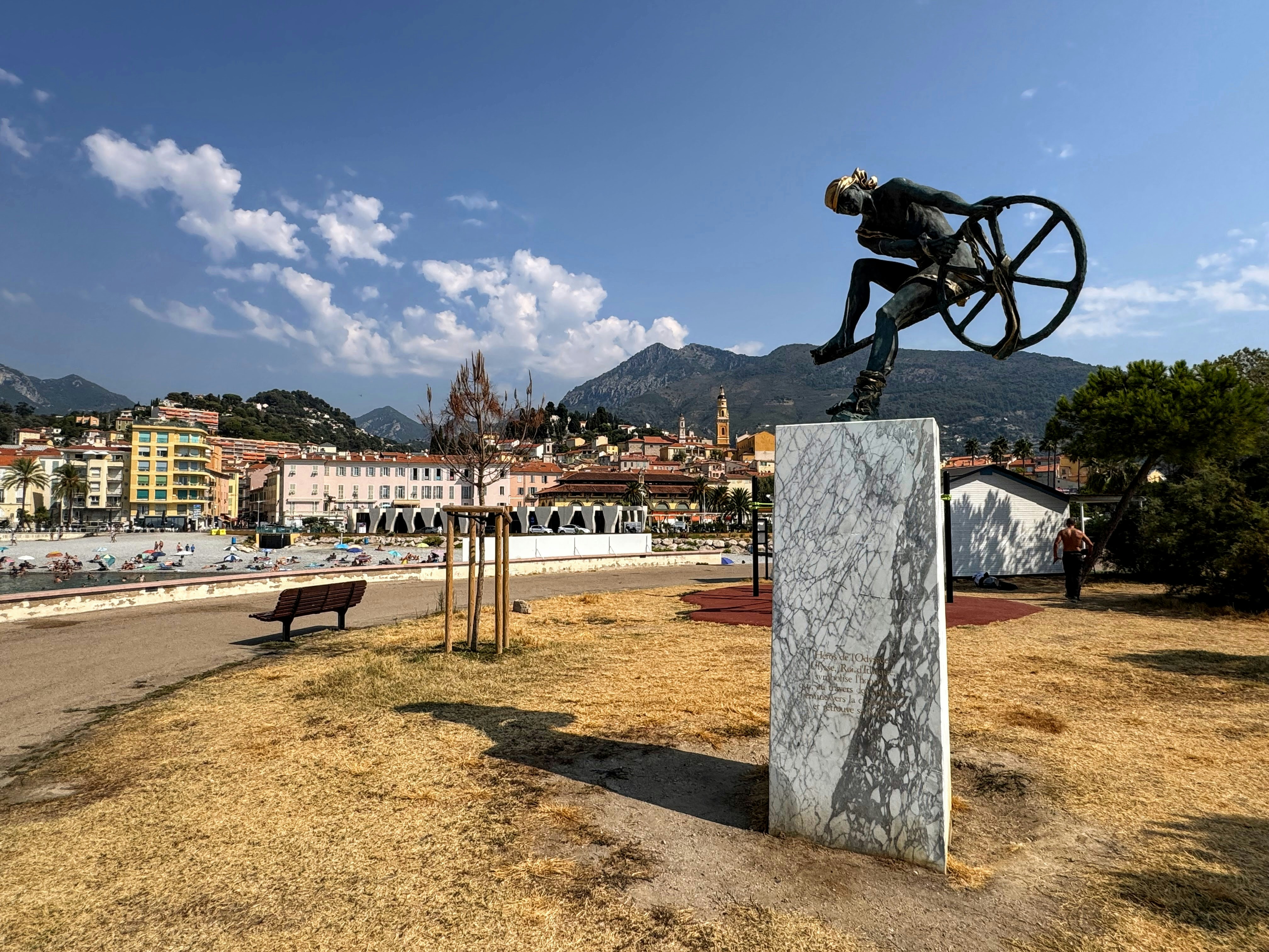 Sculpture of cyclist on pedestal with coastal town background.