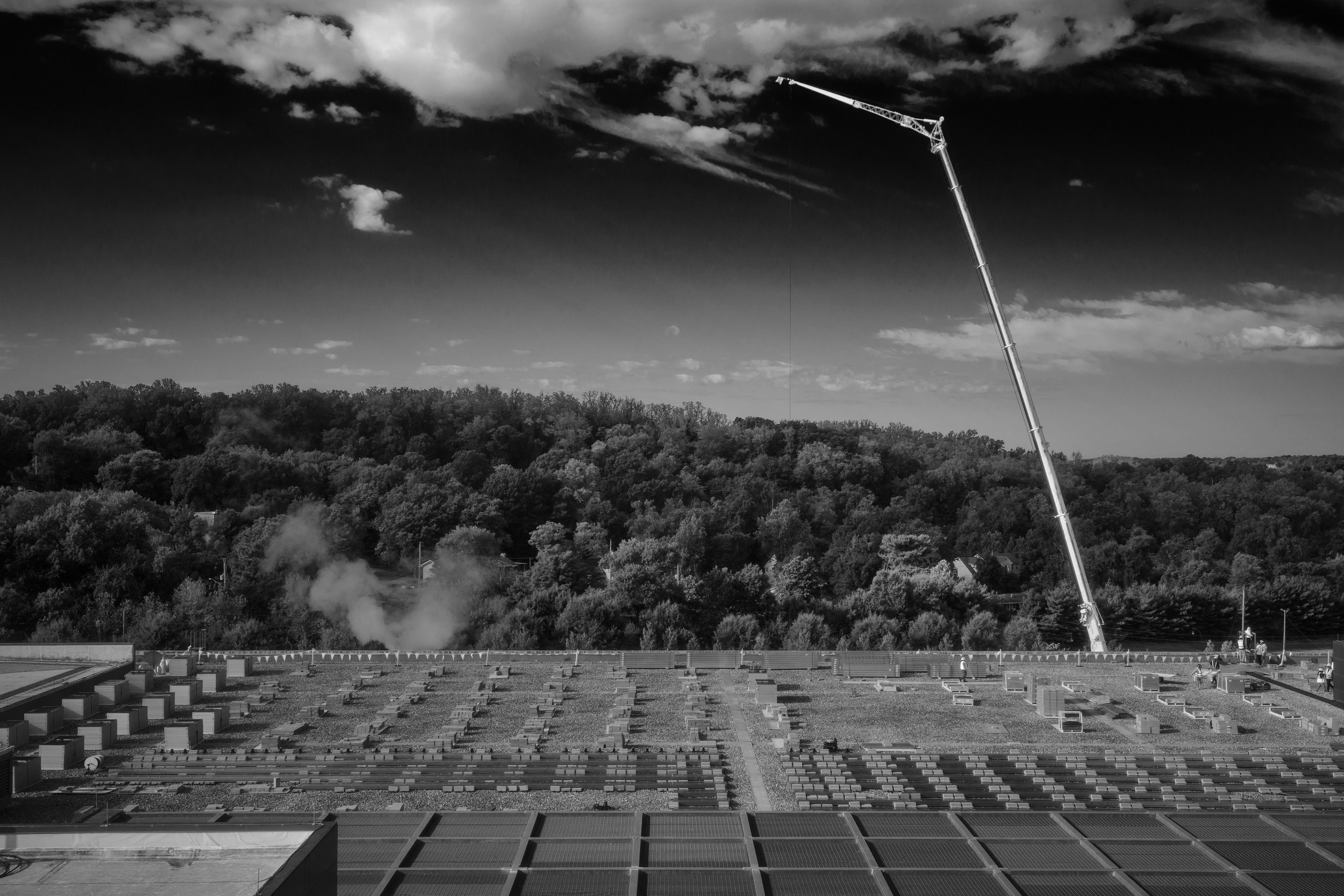 A construction crane towers over a development site against a backdrop of trees and hills. | Construction crane on a rooftop with trees behind.
