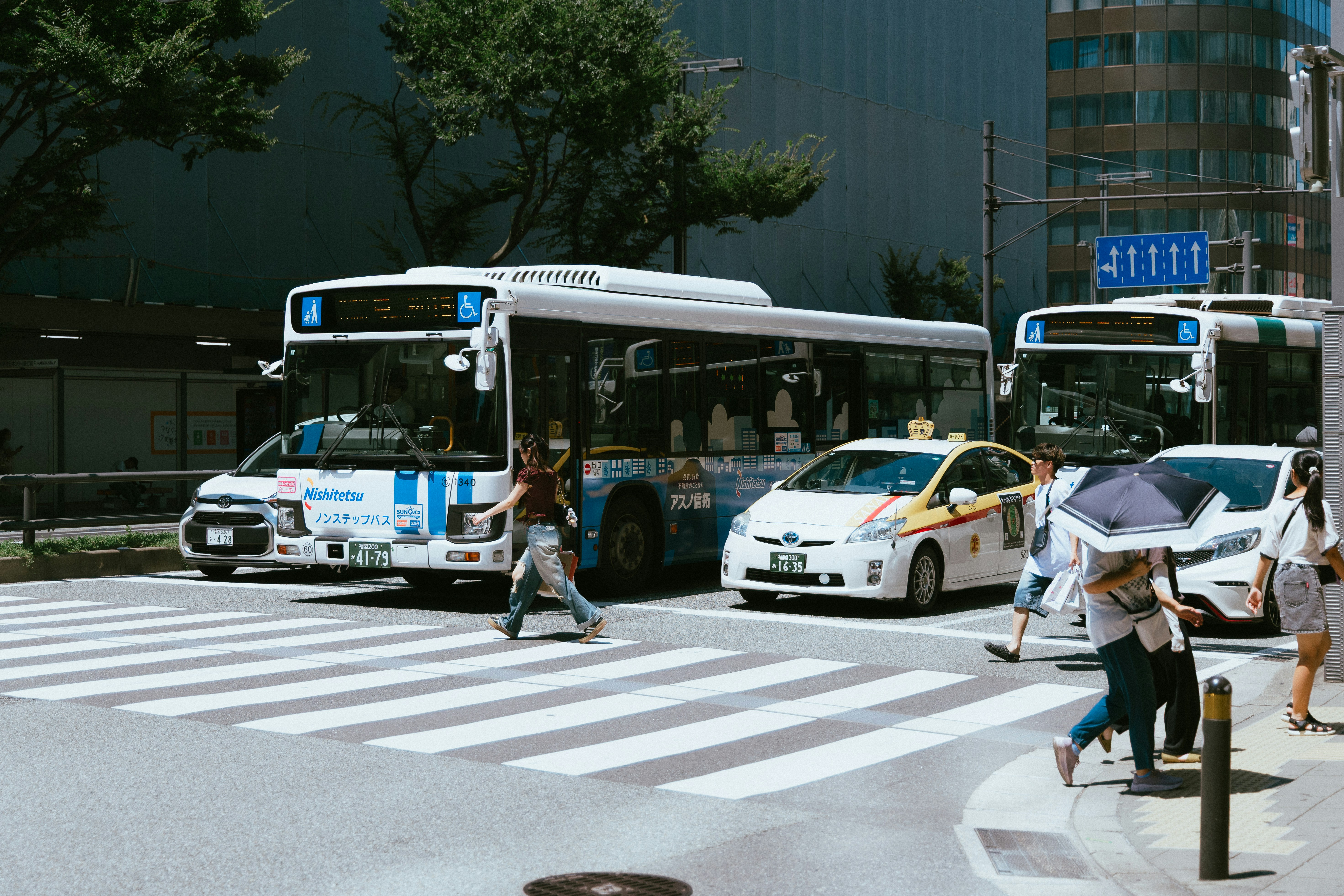 City bus and taxi navigate a bustling crosswalk as pedestrians move through a sunlit urban landscape.
