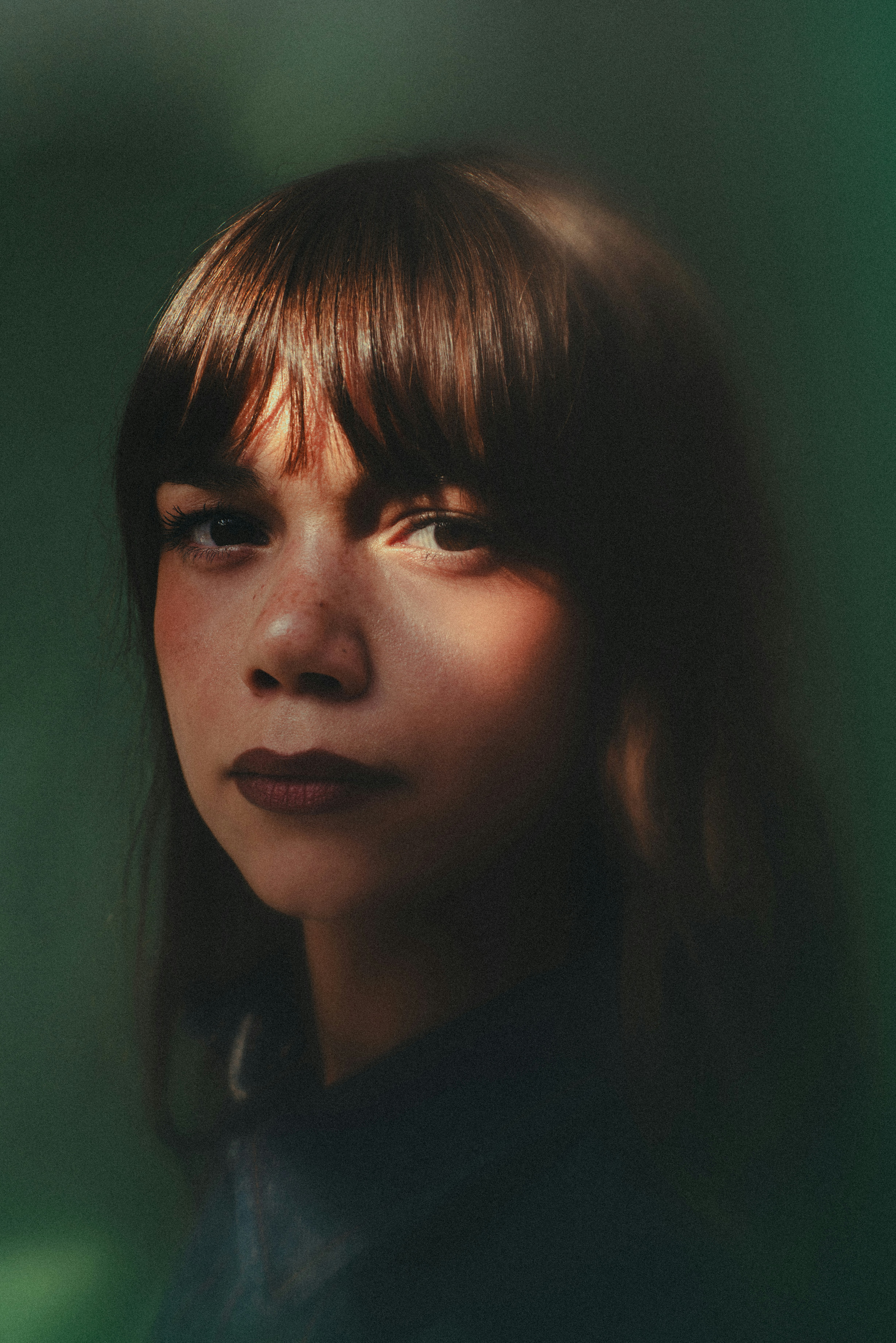 A close-up portrait featuring a young woman with soft lighting and a muted green background, highlighting her expressive features and subtle emotions.