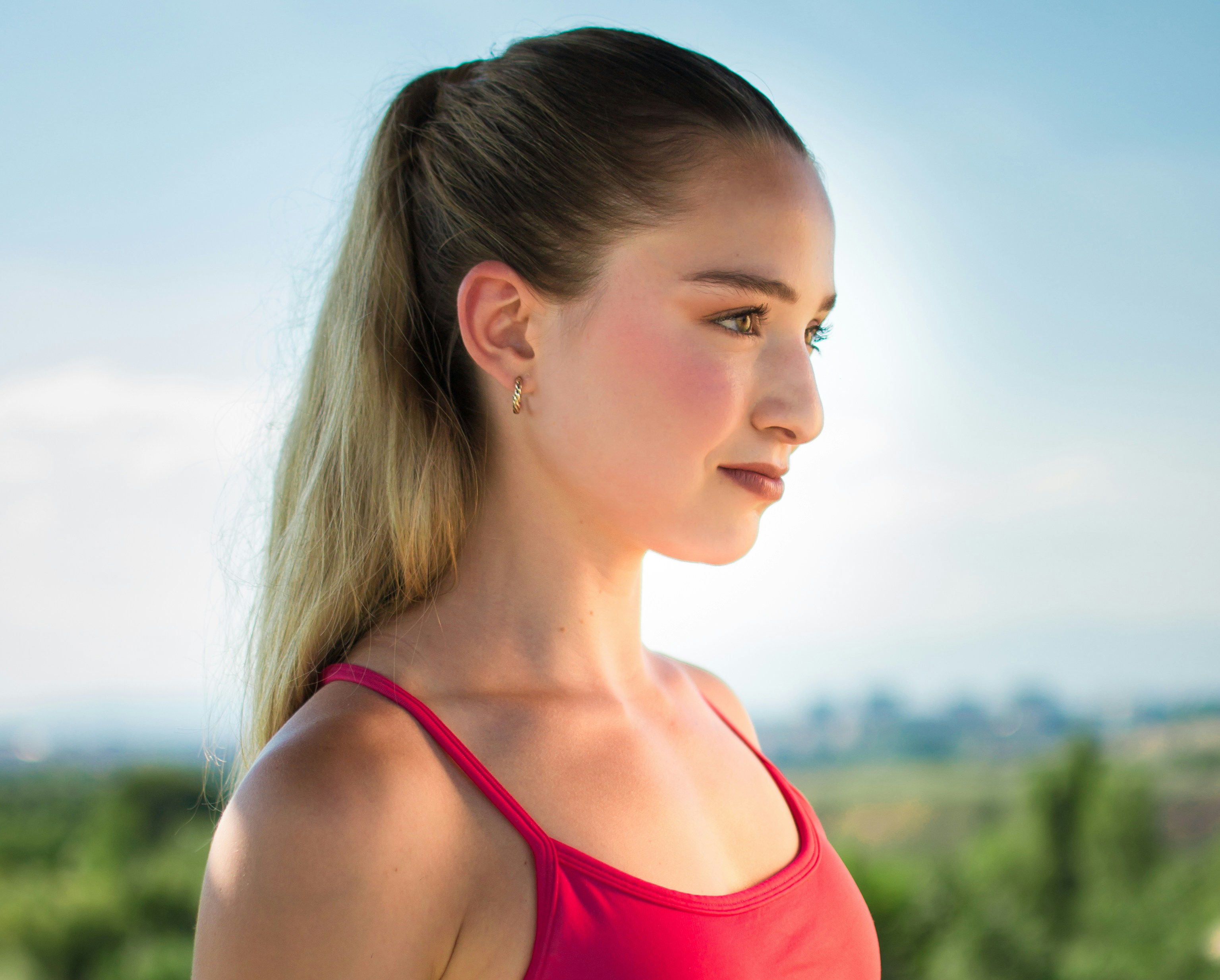 Profile of a young woman with long hair, wearing a red tank top, set against a blurred natural background. The sunlight highlights her features.