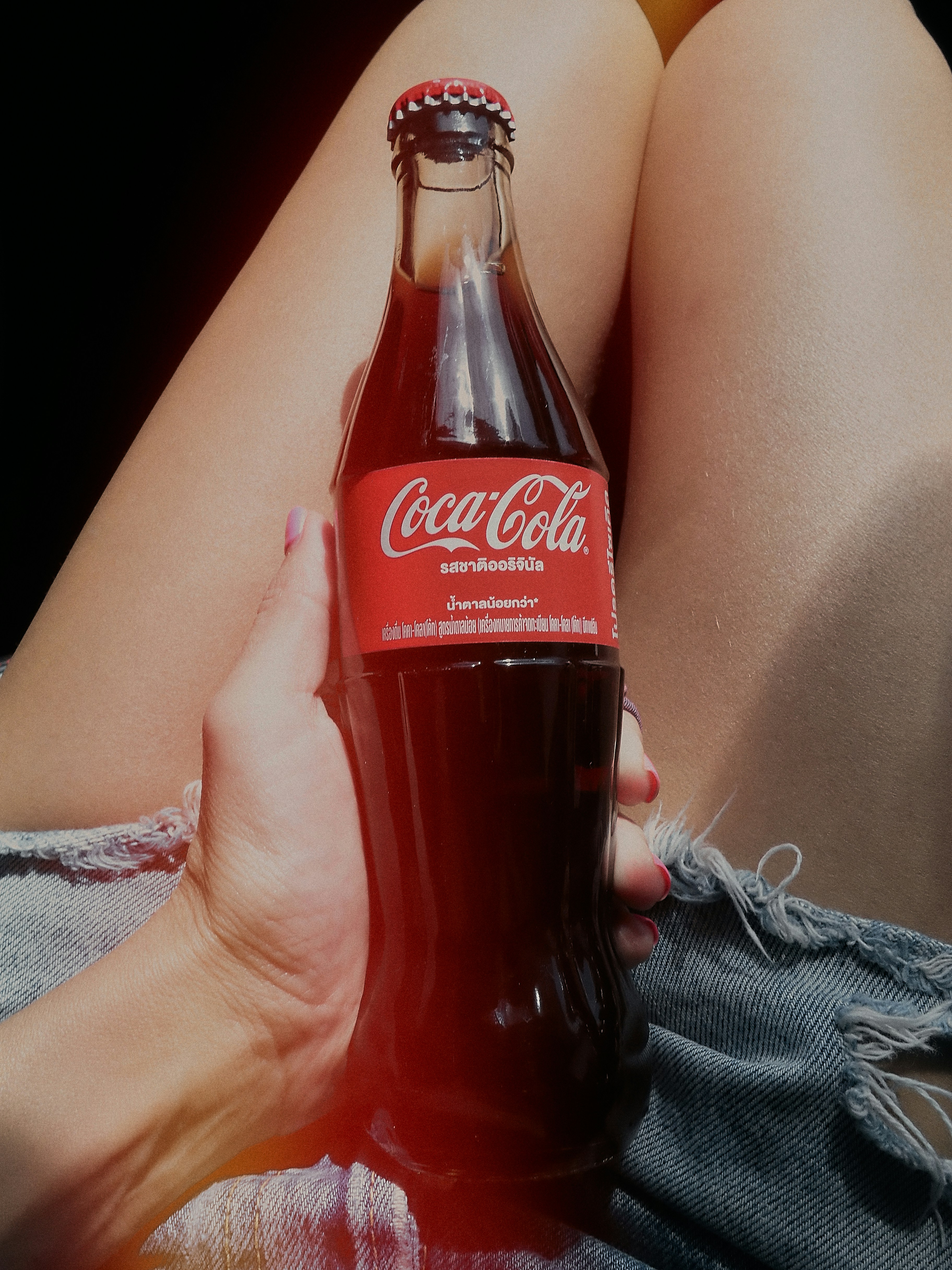A close-up of a hand holding a classic glass bottle of Coca-Cola against sun-kissed skin and ripped denim shorts. Shot in Thailand, this image captures a nostalgic summer vibe — warmth, road trips, and the simple pleasure of a cold drink. | Hand holding a coca-cola bottle between legs