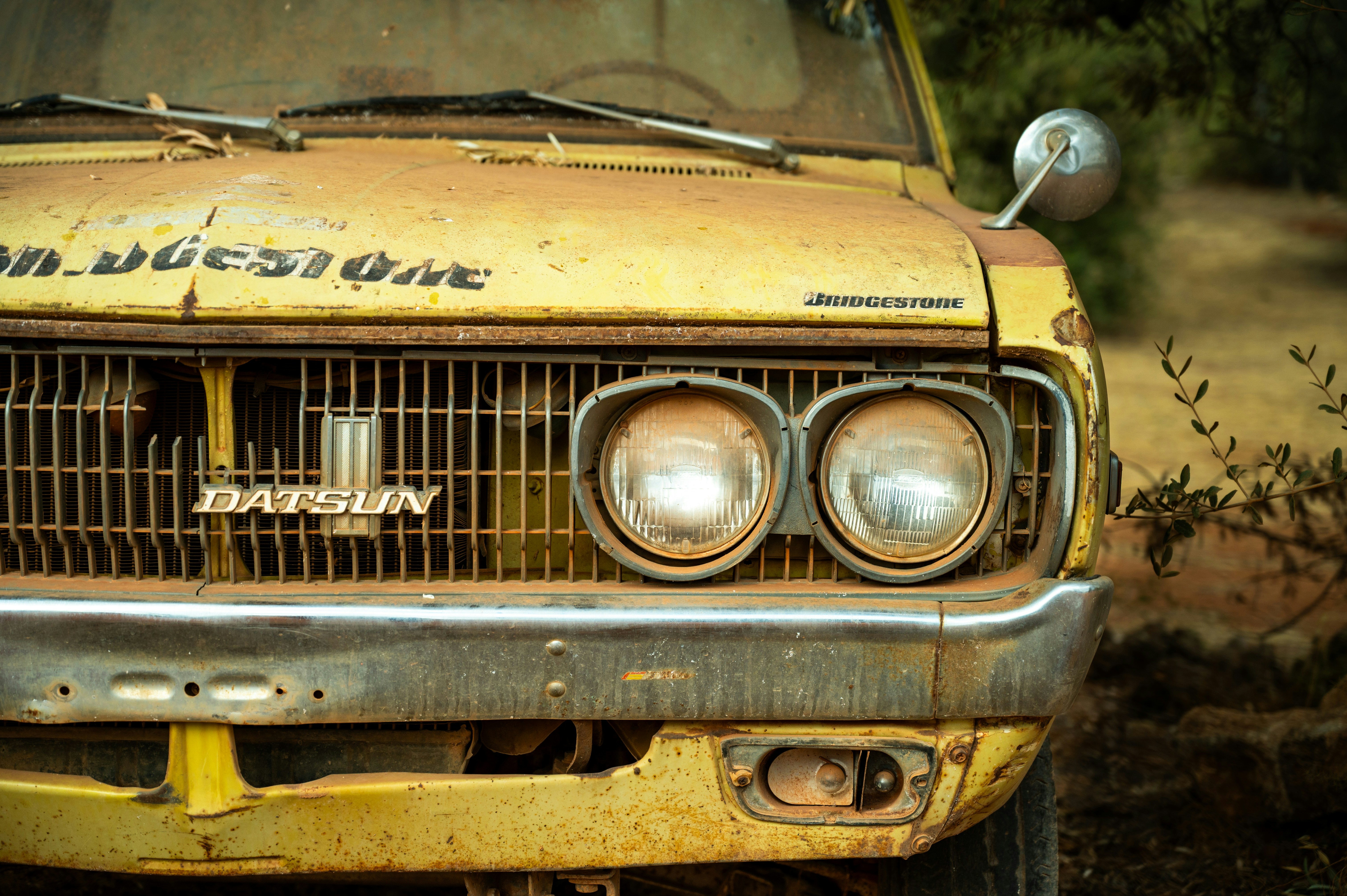 Front view of a weathered yellow Datsun, showcasing its vintage design and faded branding. The car's headlights and rusted features tell a story of time and neglect.