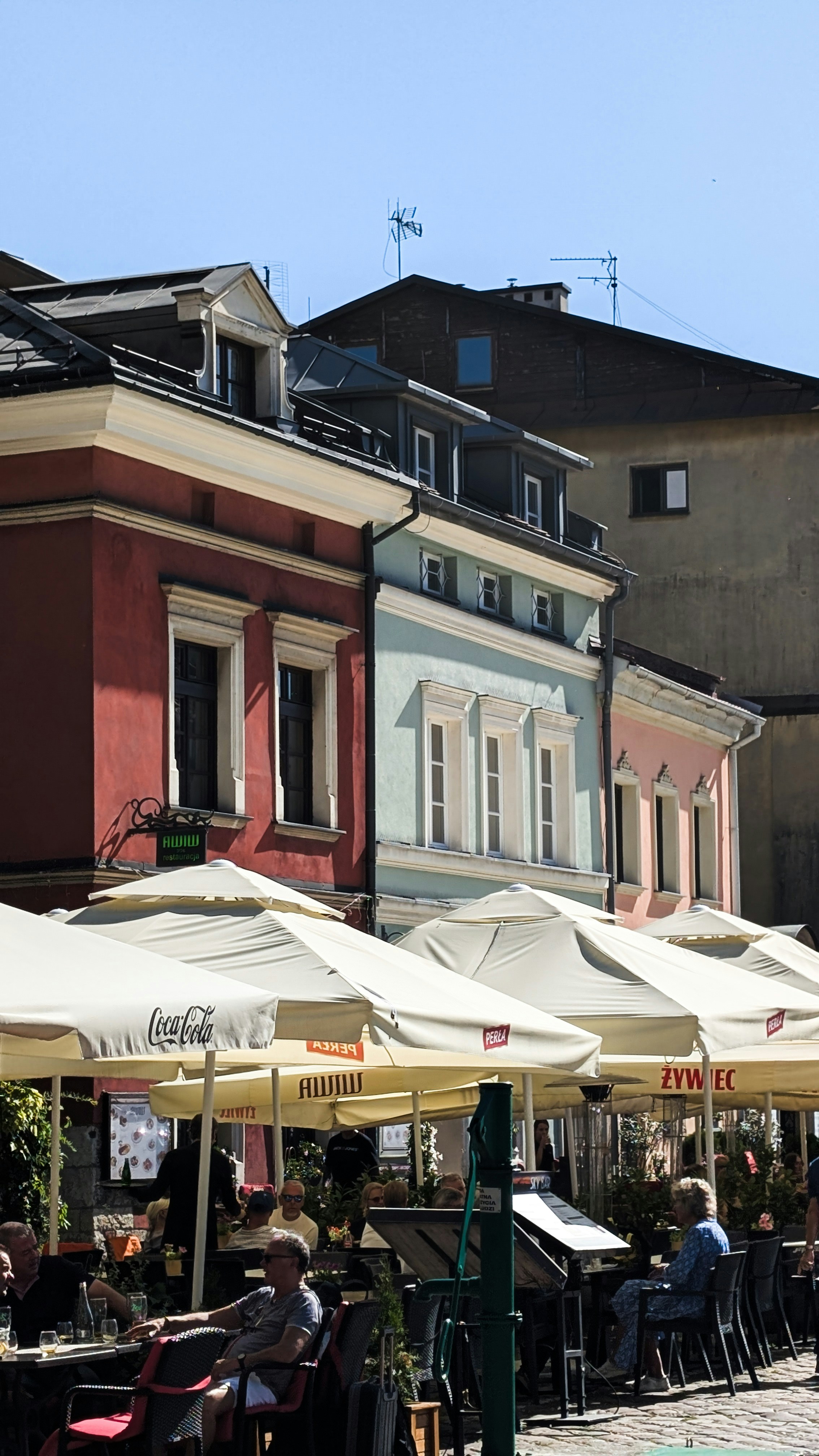 Colorful buildings frame a bustling outdoor café scene filled with patrons enjoying their meals under large umbrellas.