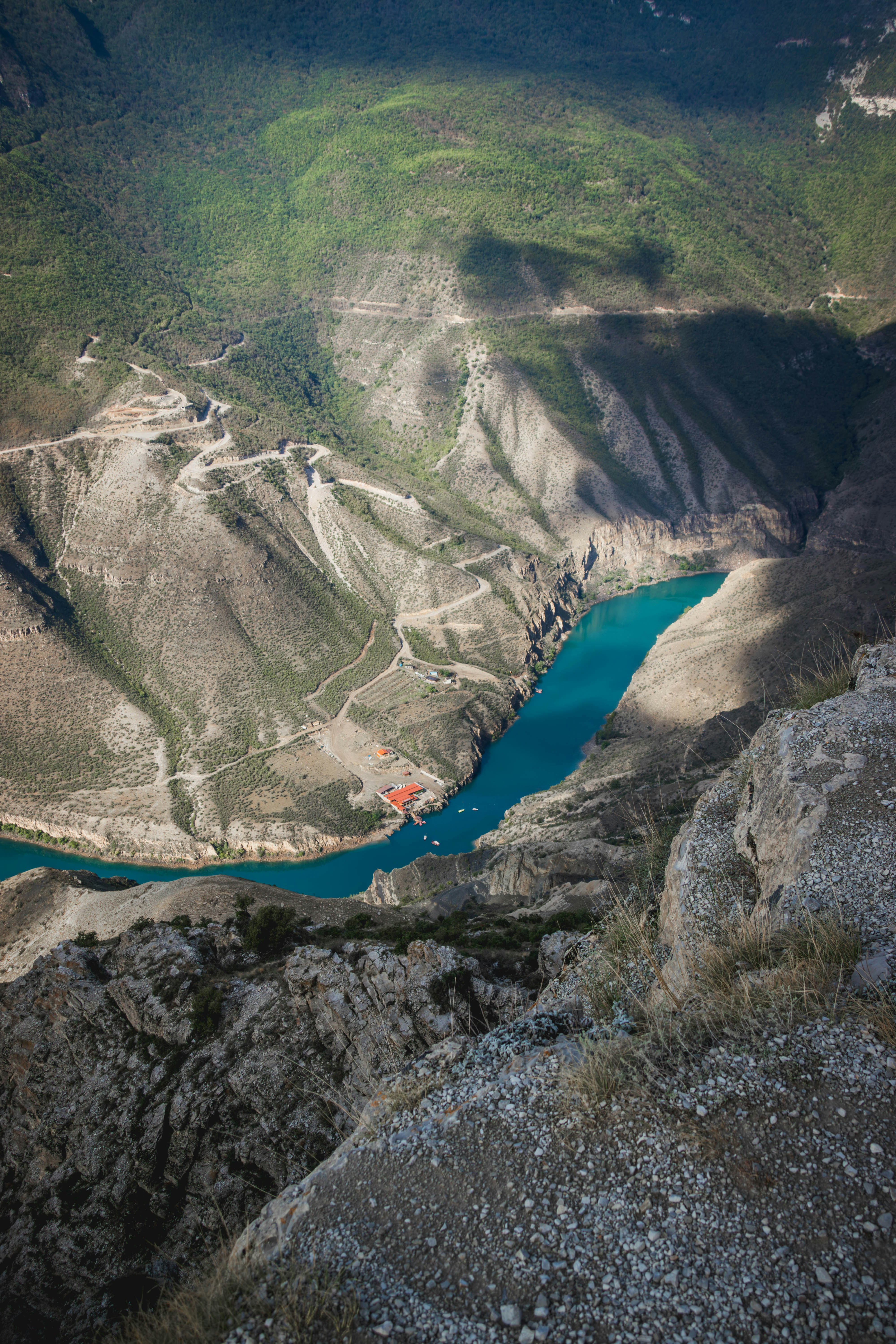 Aerial view of a vibrant blue river winding through rugged terrain, flanked by steep cliffs and patches of greenery. The scene captures the interplay of light and shadow on the landscape.