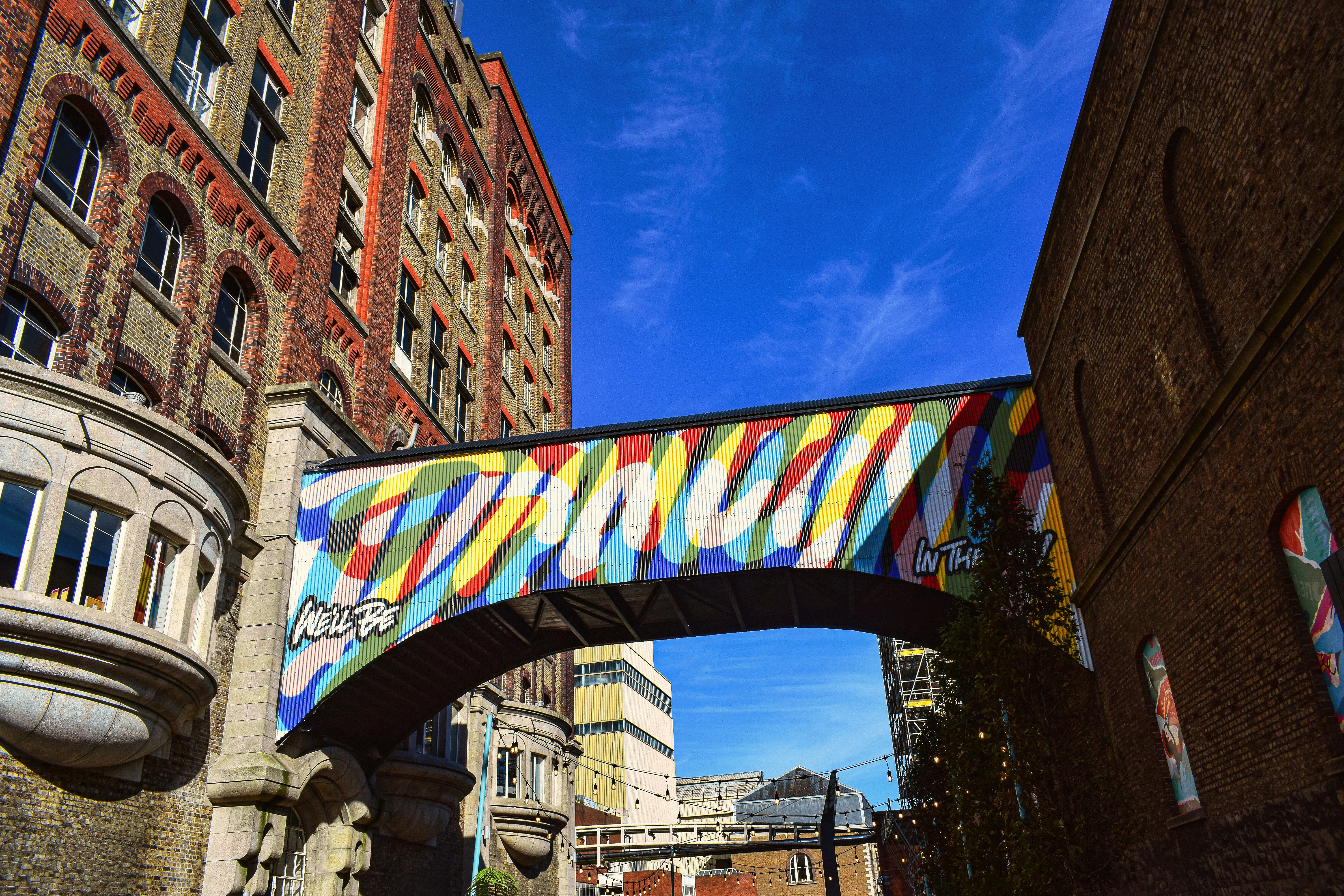 Colorful mural adorning a bridge between historic buildings, showcasing urban art against a clear blue sky.