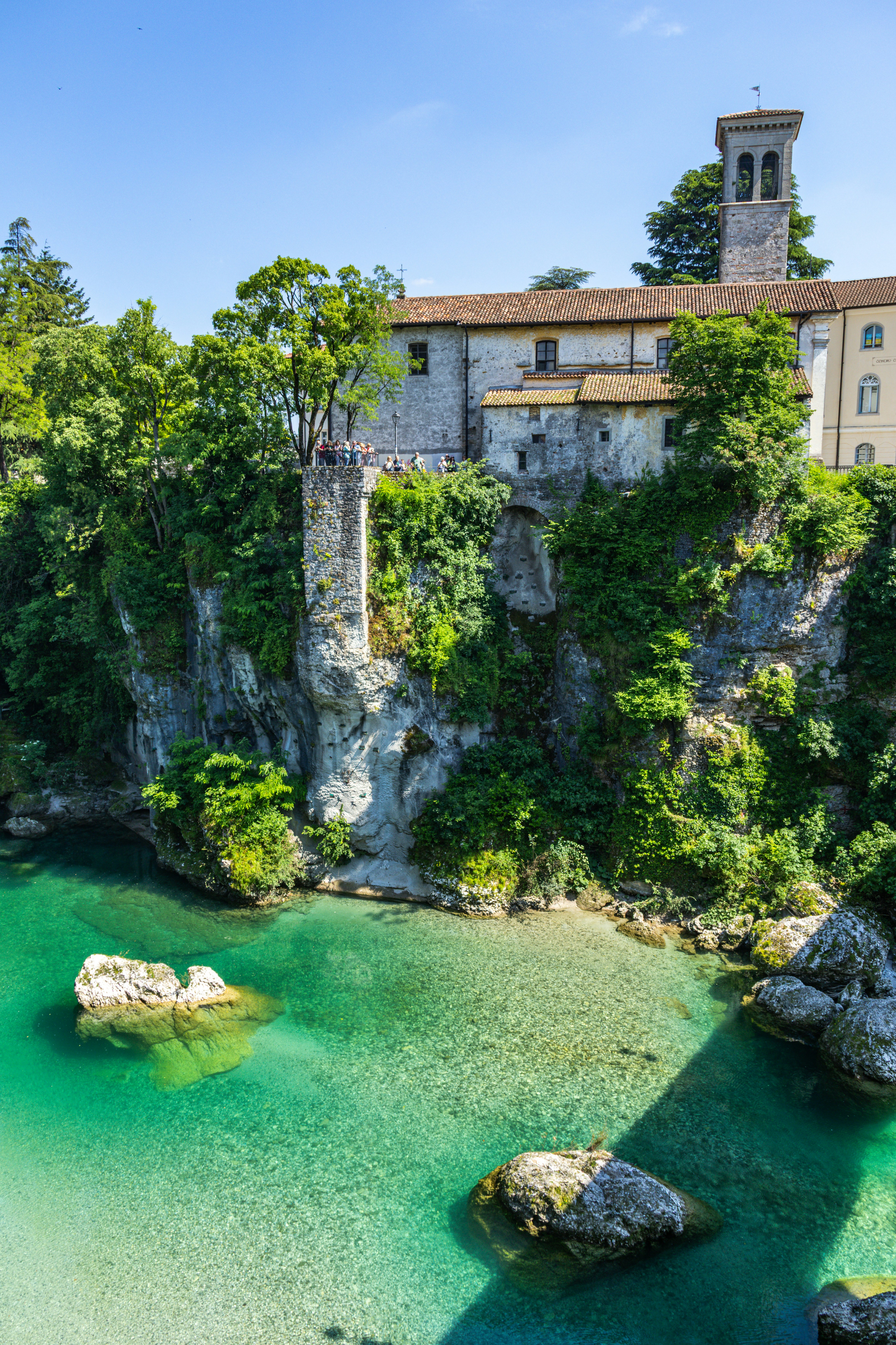 A historic structure perched on a rocky cliff, surrounded by lush greenery and reflecting in the tranquil turquoise waters below.