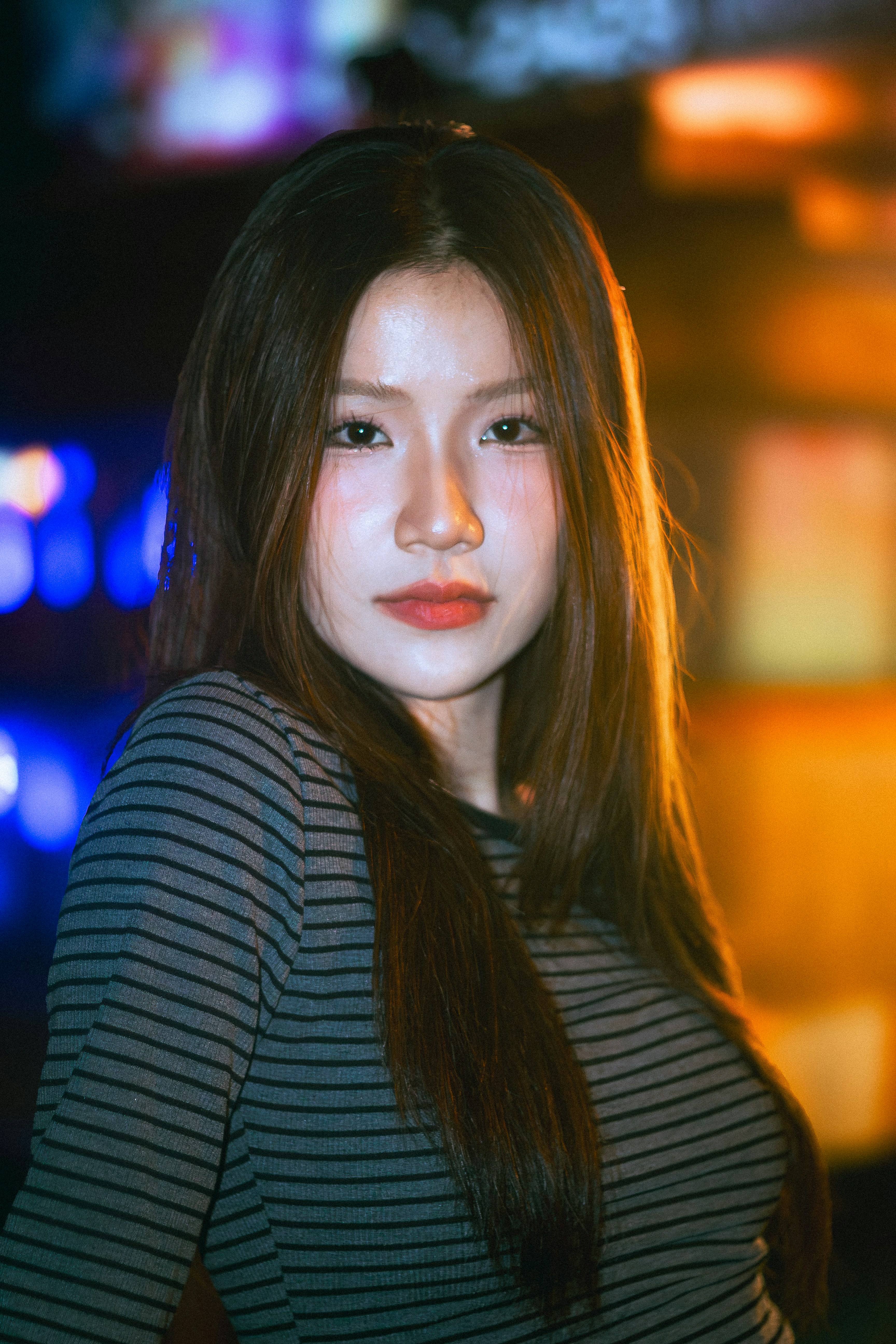 A young woman with long hair poses confidently against a backdrop of colorful lights, showcasing her striking features and relaxed demeanor.