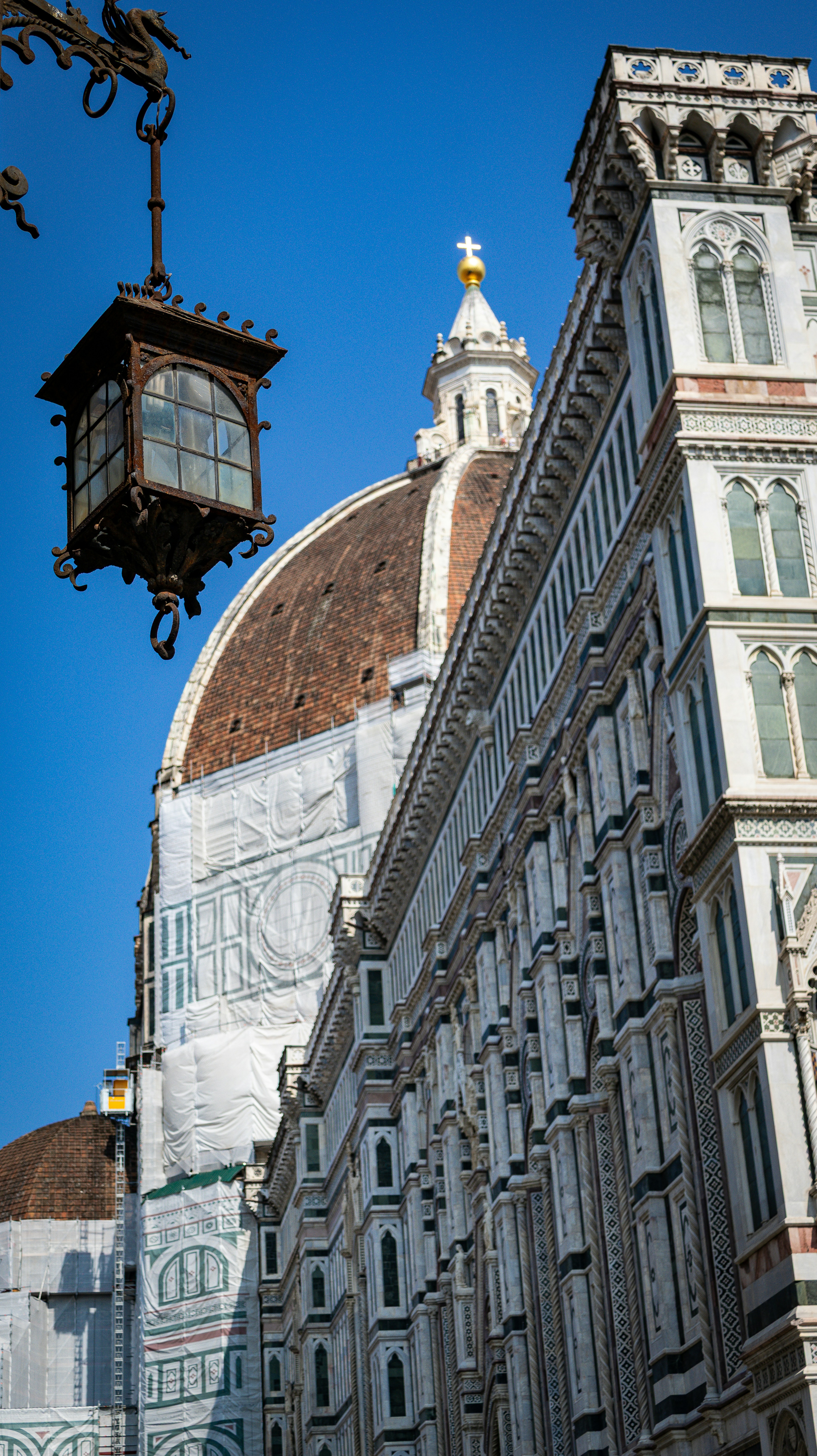 Historic cathedral dome shrouded in scaffolding, juxtaposed with a vintage lantern under a clear blue sky.
