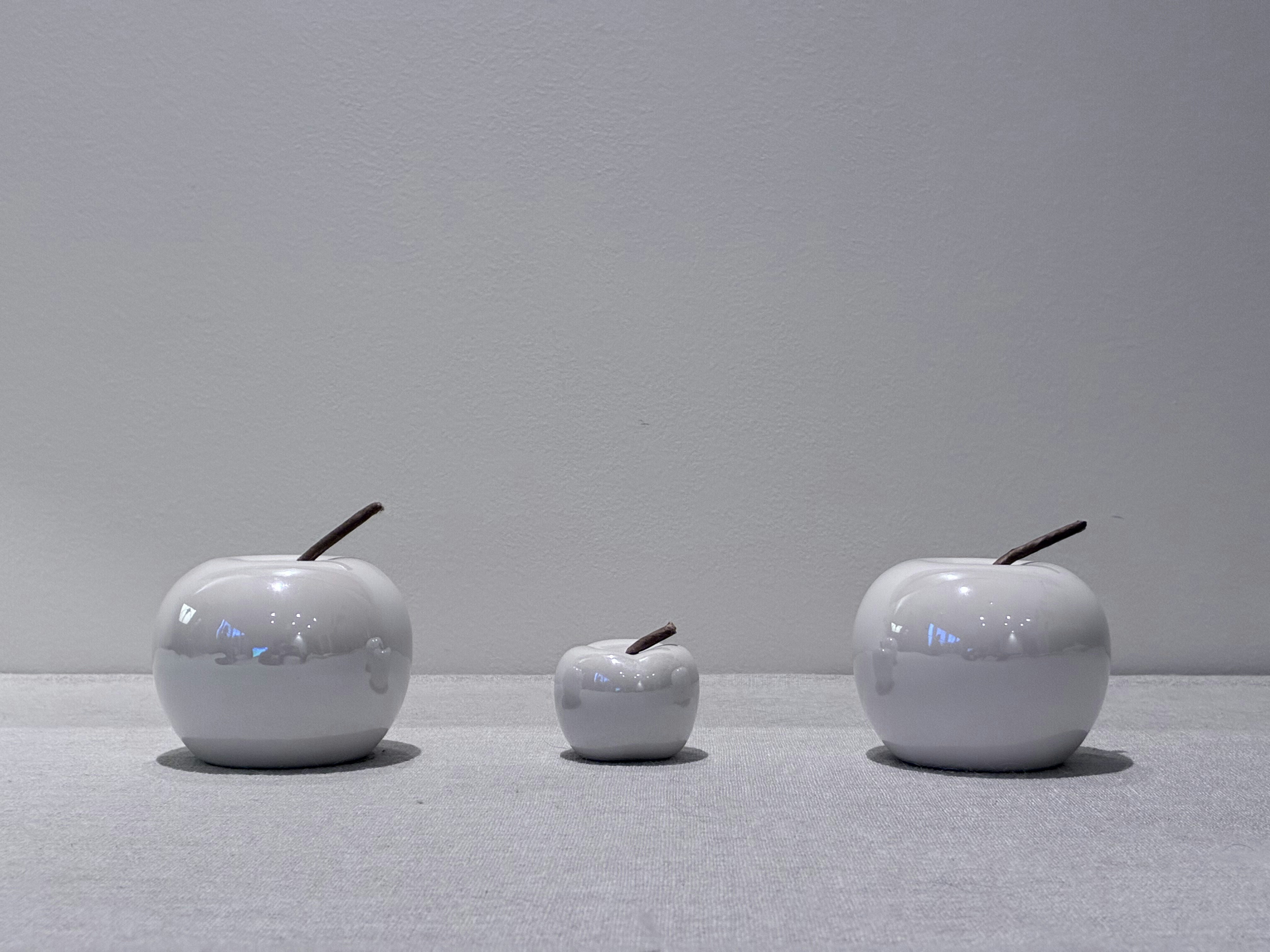 Three porcelain apple sculptures of varying sizes arranged on a neutral backdrop, showcasing their smooth surfaces and subtle reflections.