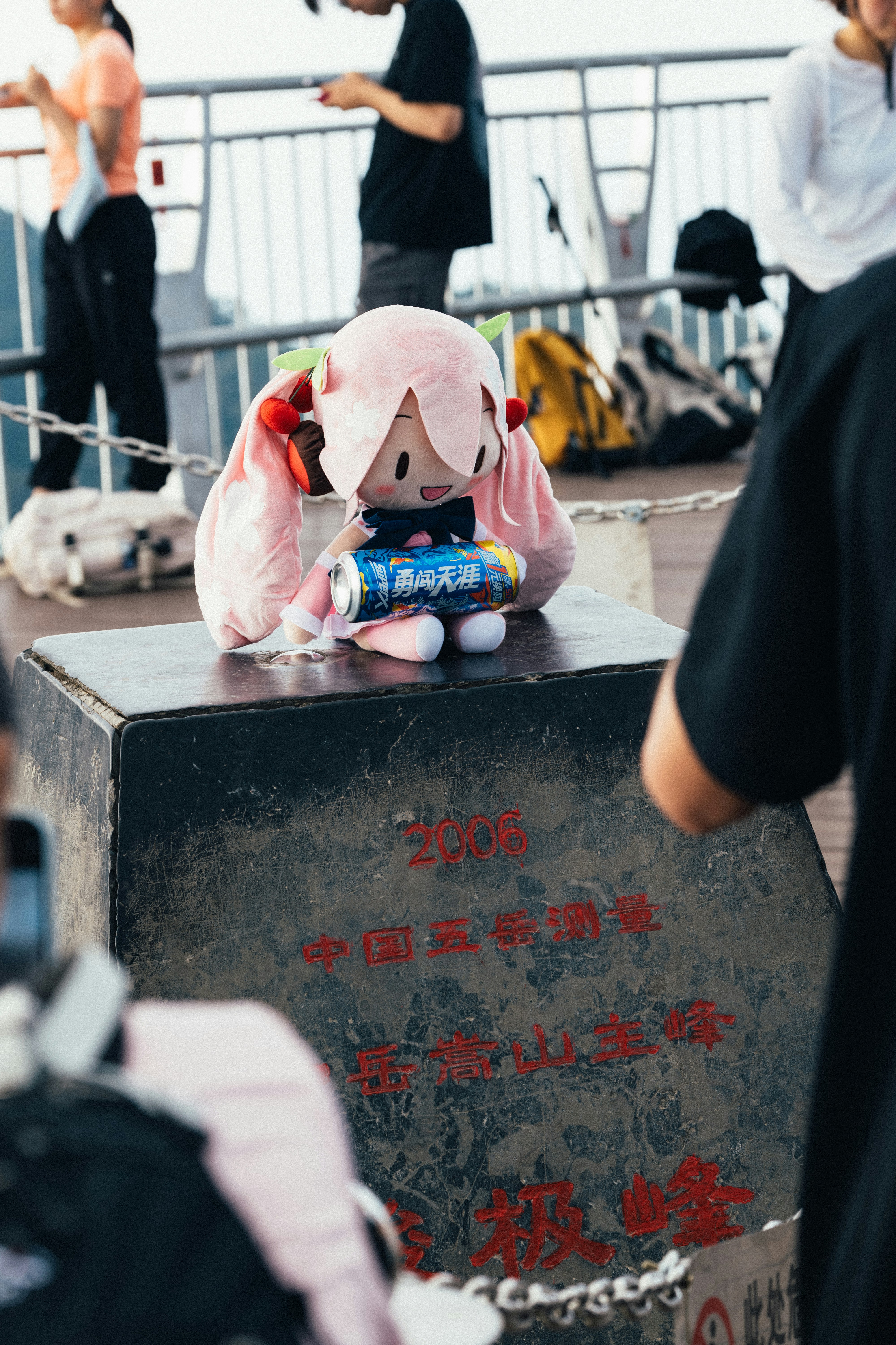 Plush doll sits on a stone monument with people around.