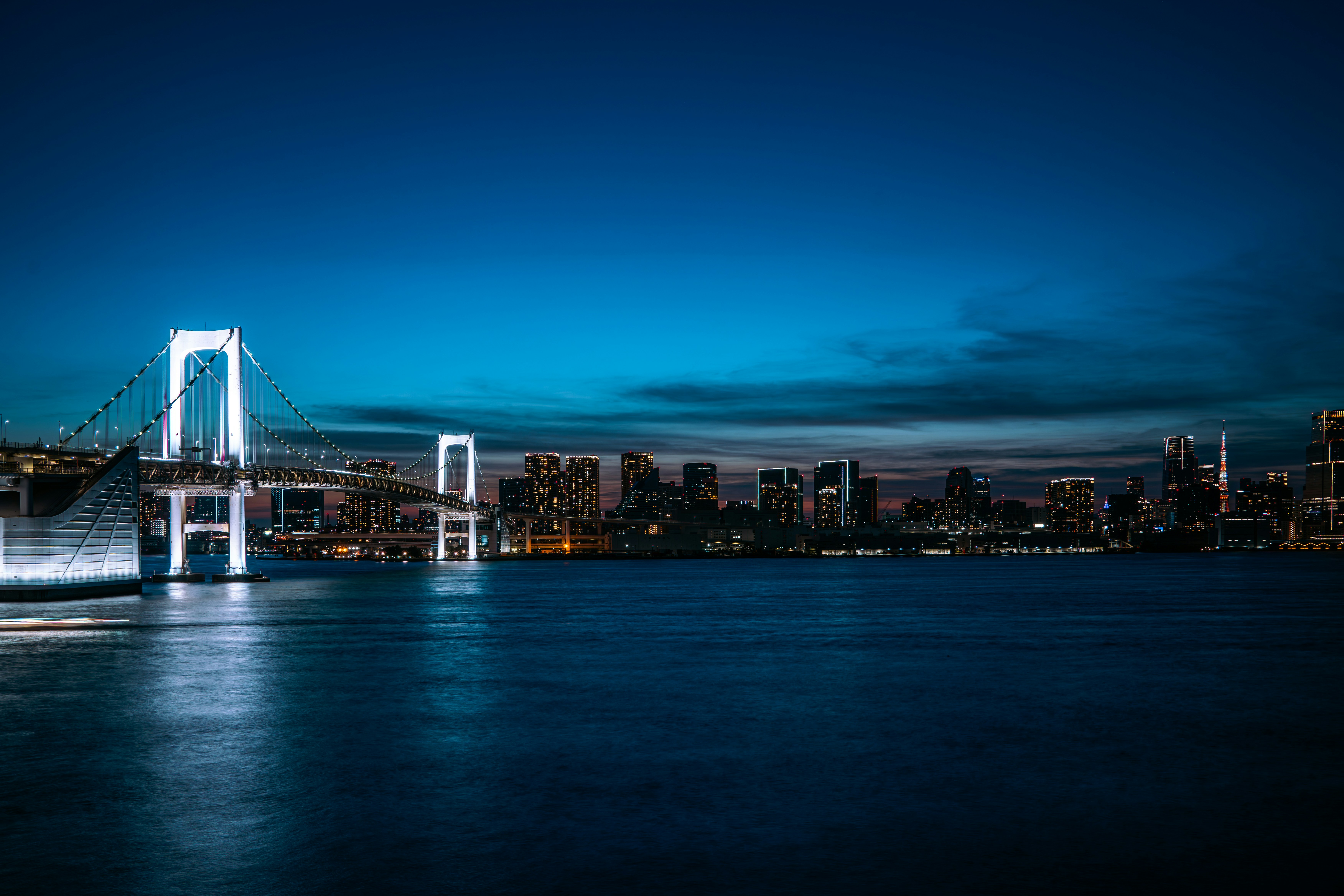 Illuminated bridge spanning a tranquil bay, with a vibrant city skyline reflecting on the water's surface at dusk.