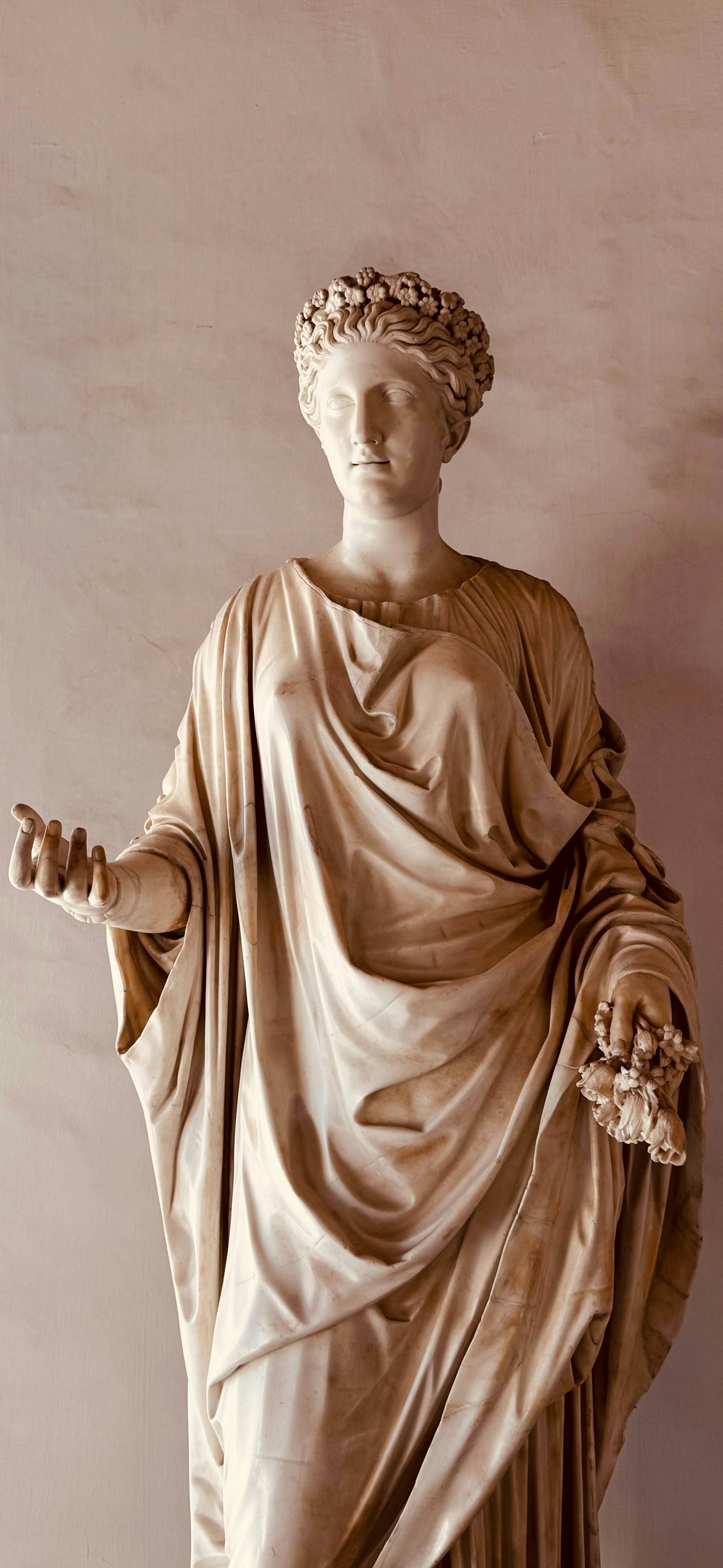 A marble statue depicting a serene figure draped in flowing garments, holding flowers in one hand and extending the other gracefully.
