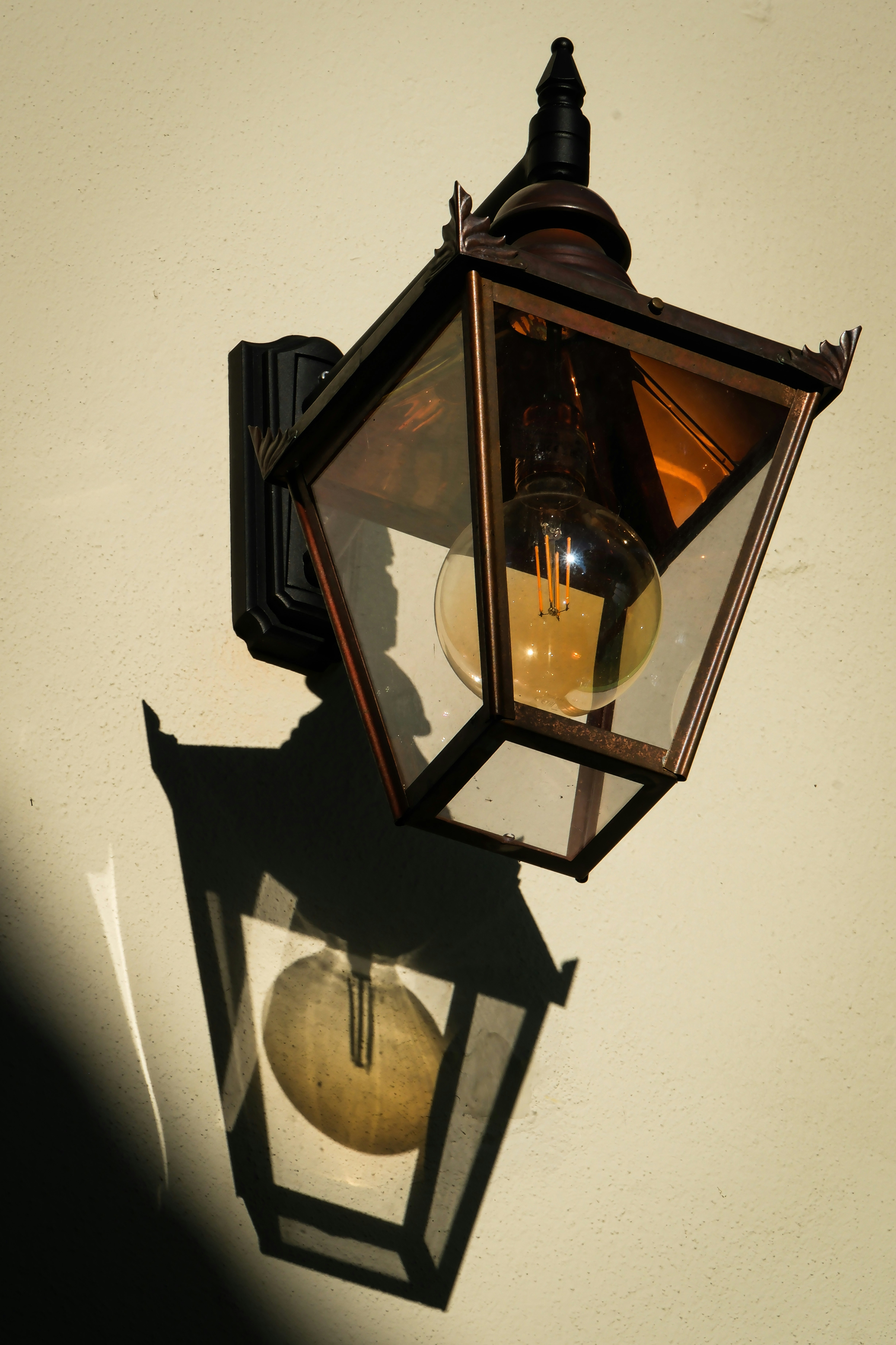 A quiet moment of light and shadow: a vintage wall lantern casts its sharp silhouette onto a pale facade. | An old-fashioned lantern casts a shadow on wall.