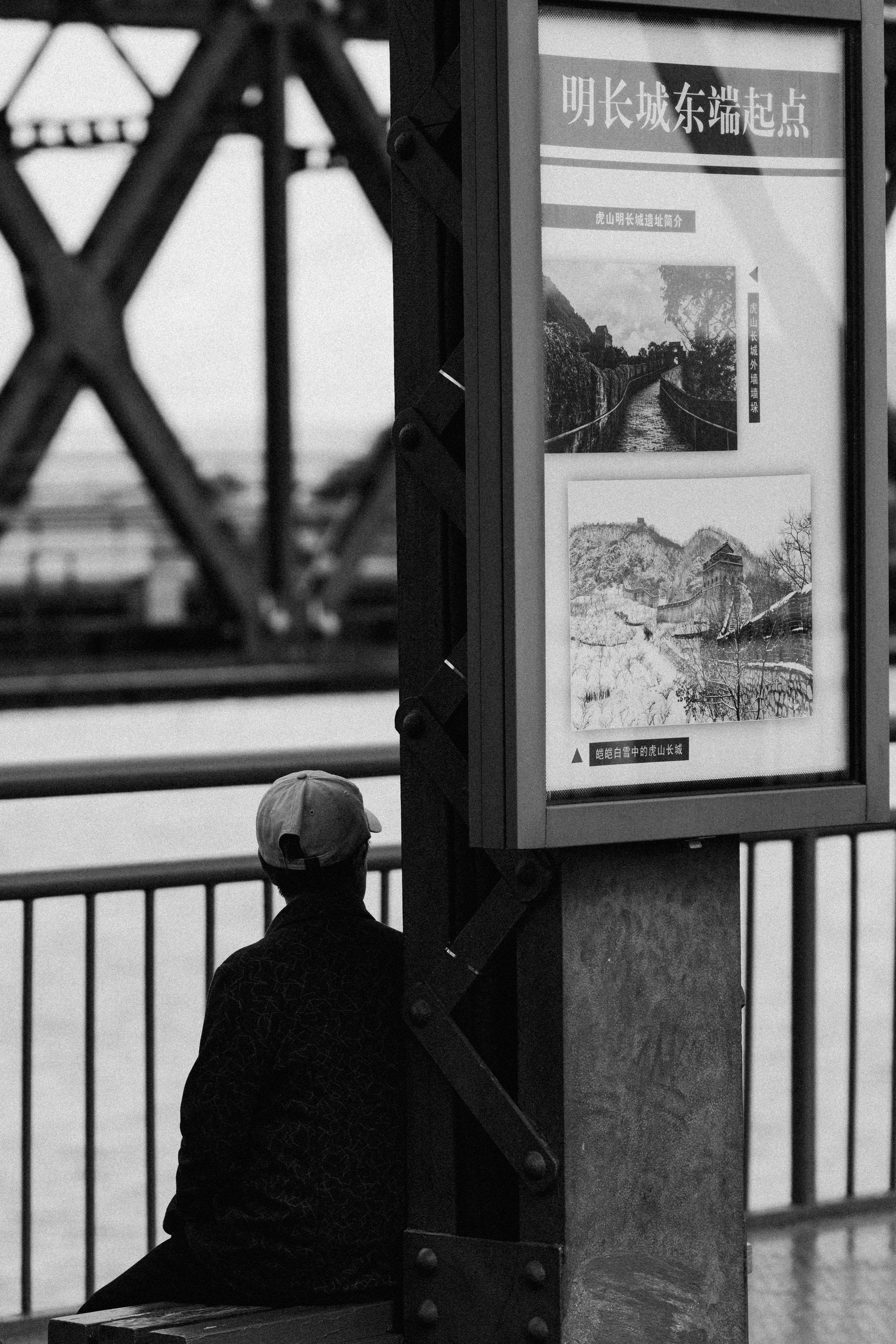 A solitary figure gazes at historical images displayed on a sign, set against a backdrop of a bridge and river. The scene evokes a sense of contemplation and connection to the past.