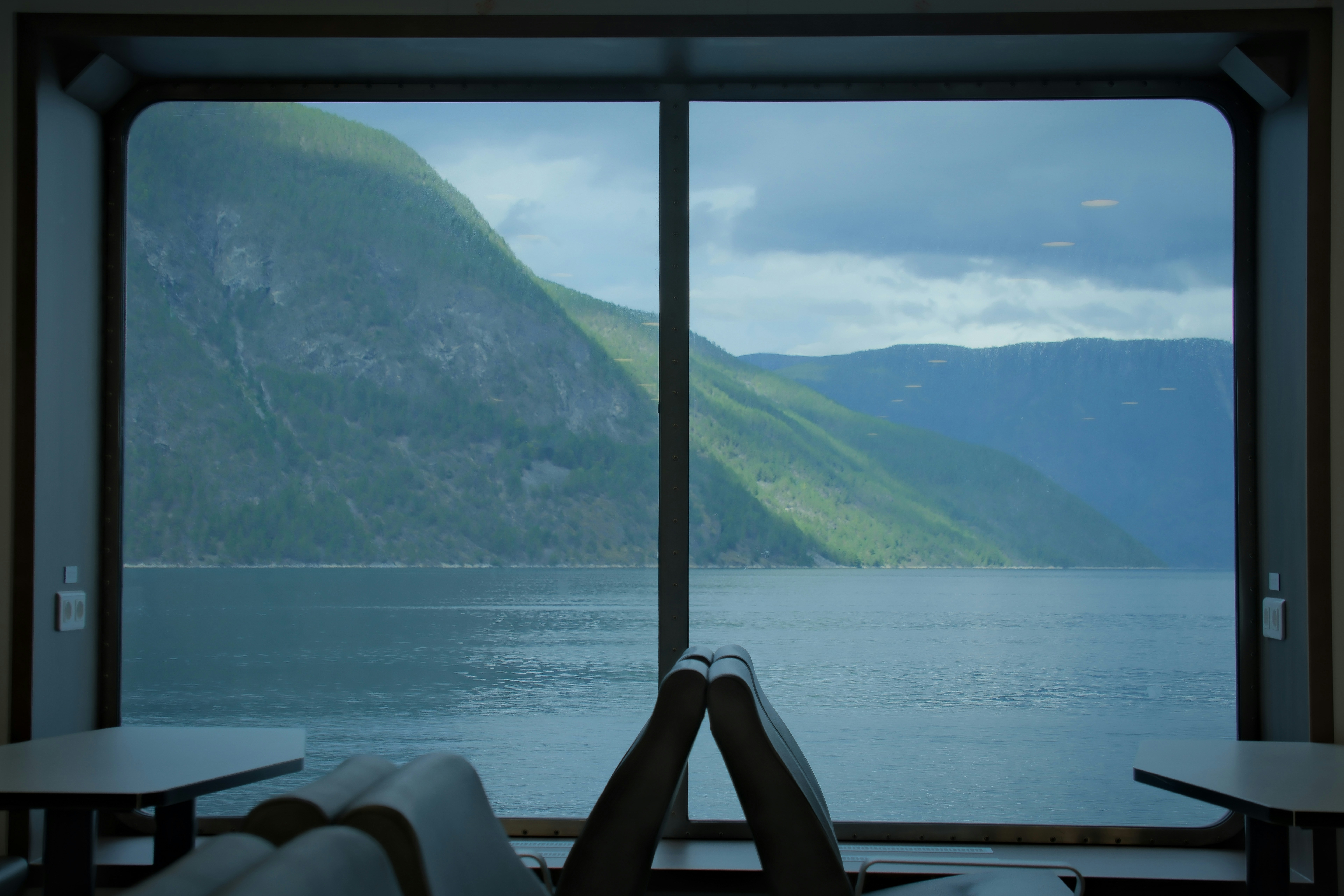 A tranquil view through large windows showcasing a fjord surrounded by lush mountains under a cloudy sky.