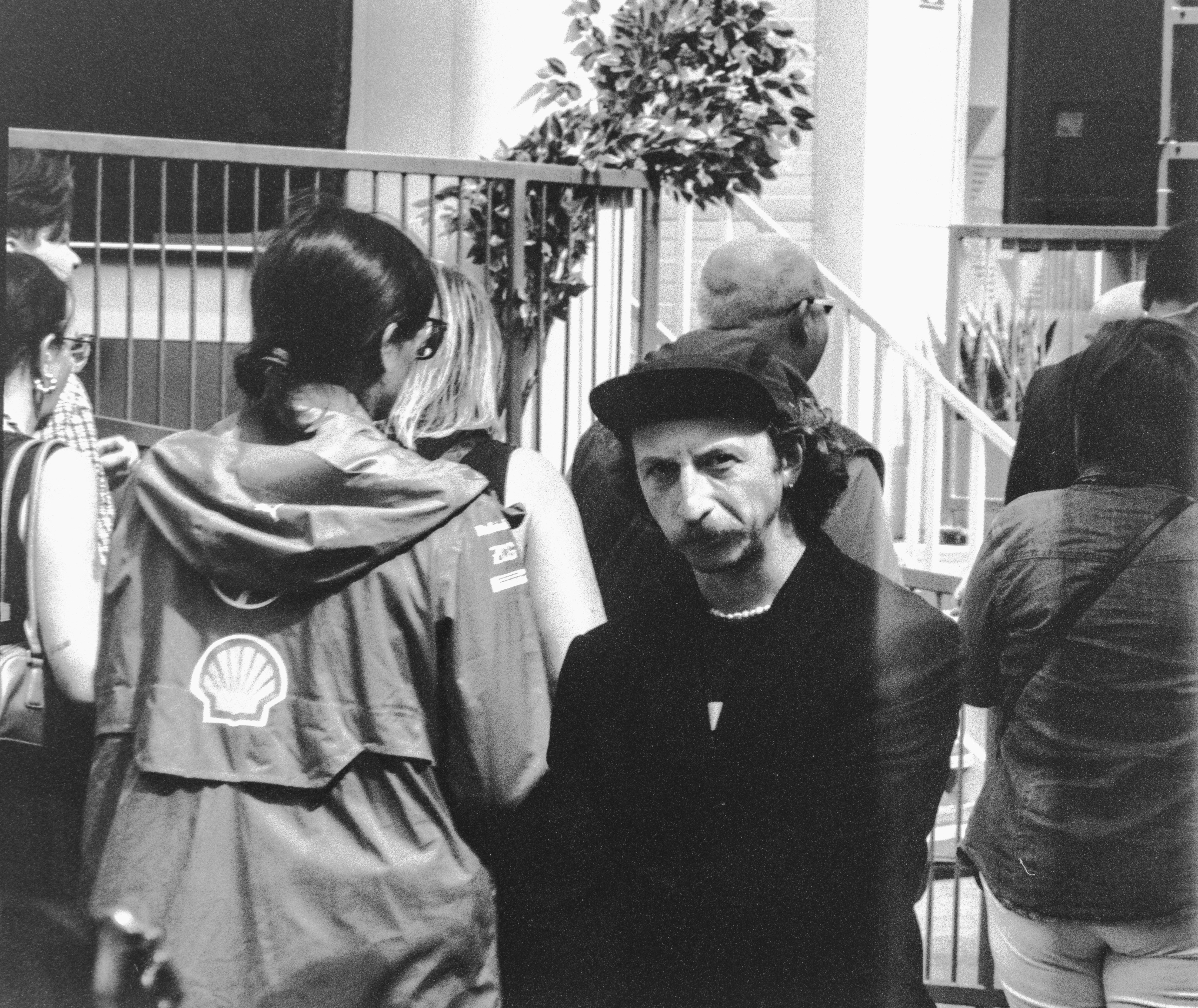 A man in a black outfit gazes intently at the camera, surrounded by a crowd of people in casual attire. The scene captures the contrast between his focused expression and the bustling background.