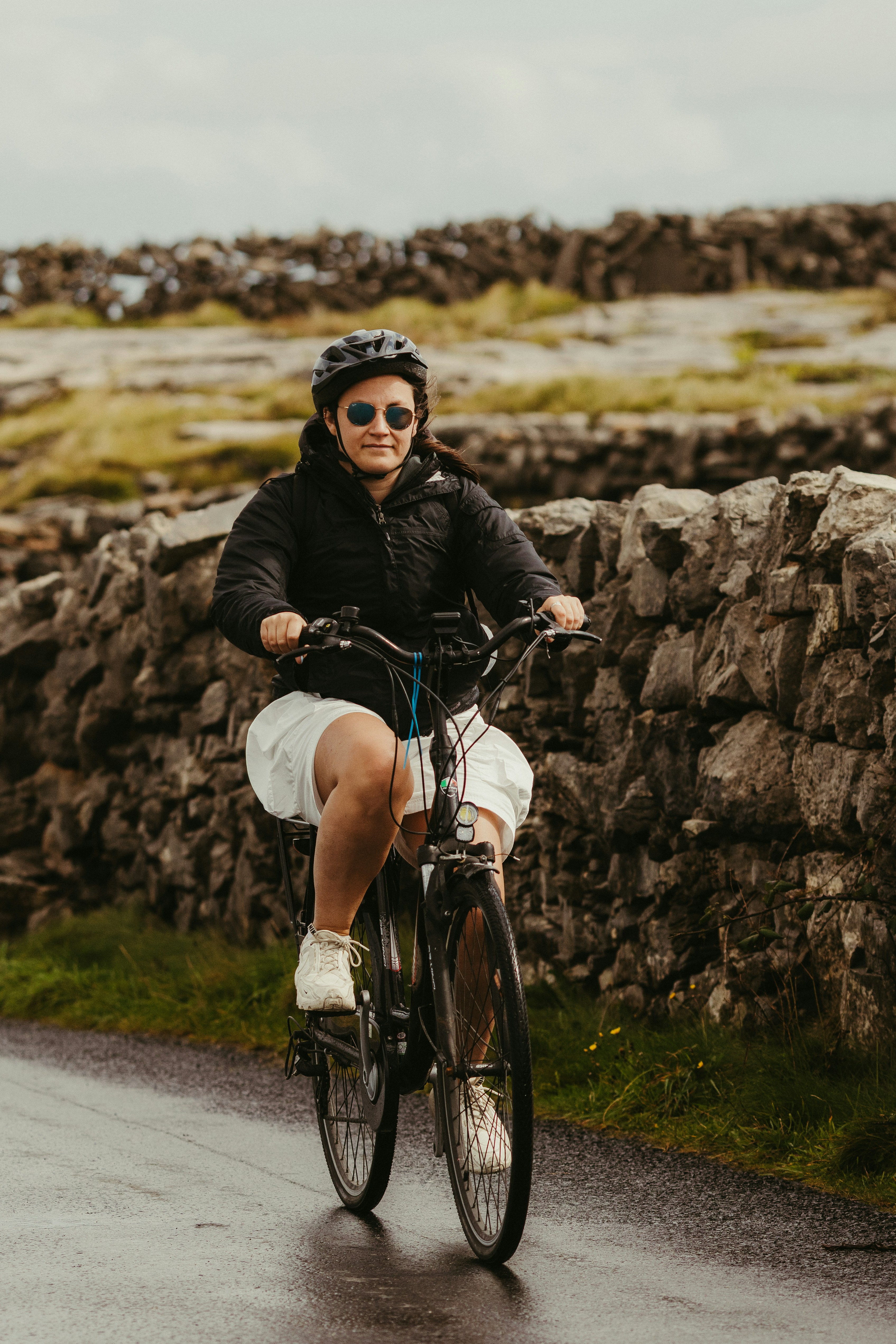 A cyclist in a black jacket and sunglasses rides along a stone wall, surrounded by lush greenery and a cloudy sky.