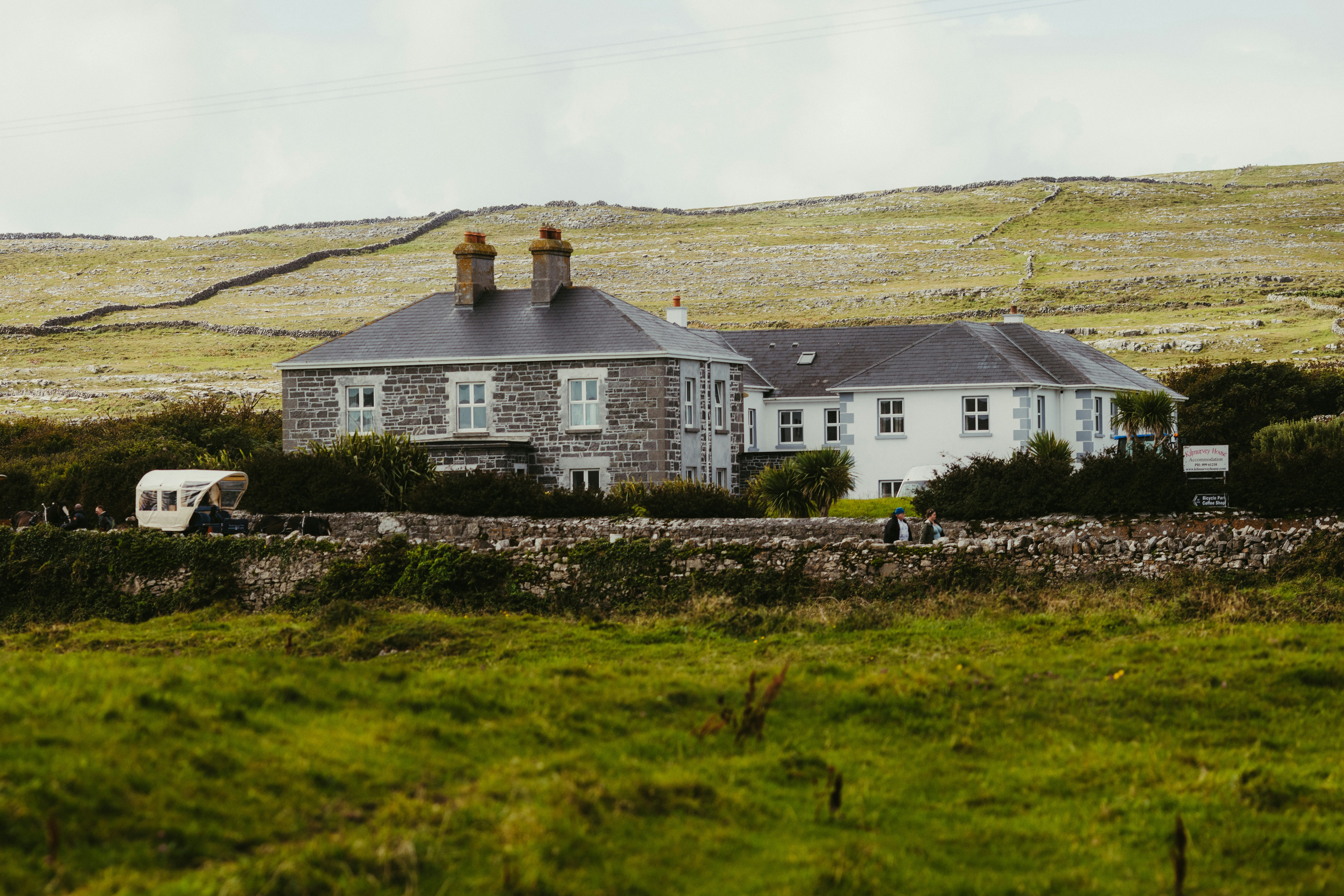 Historic stone house nestled in lush green fields, surrounded by rolling hills and a rustic wall. A glimpse of daily life unfolds nearby.