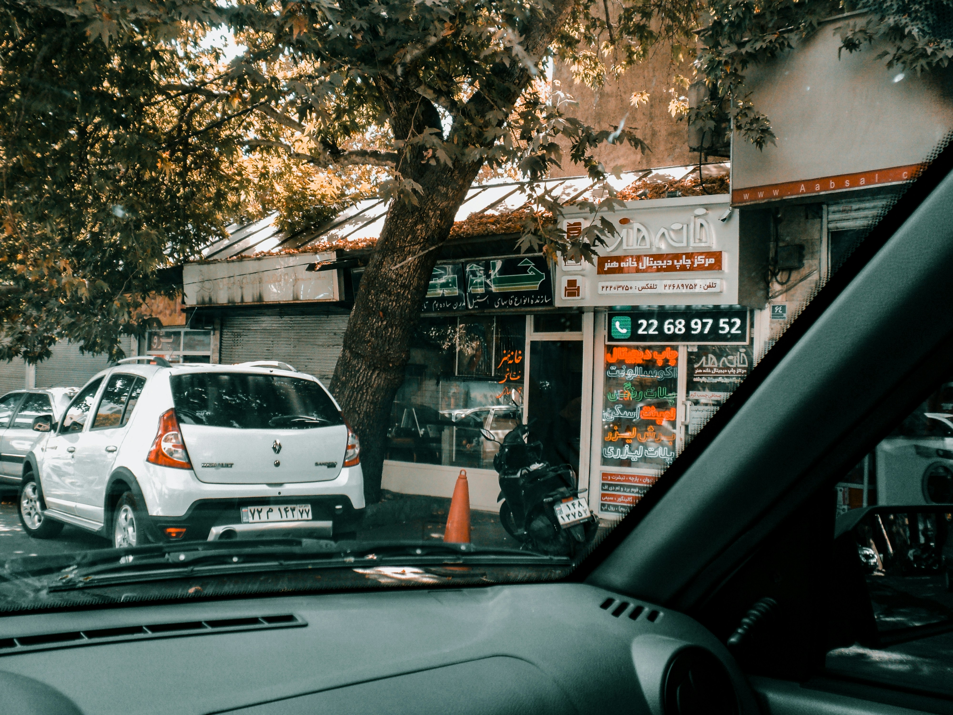 A bustling street scene featuring a food stall and parked cars under a leafy tree, showcasing everyday urban life.