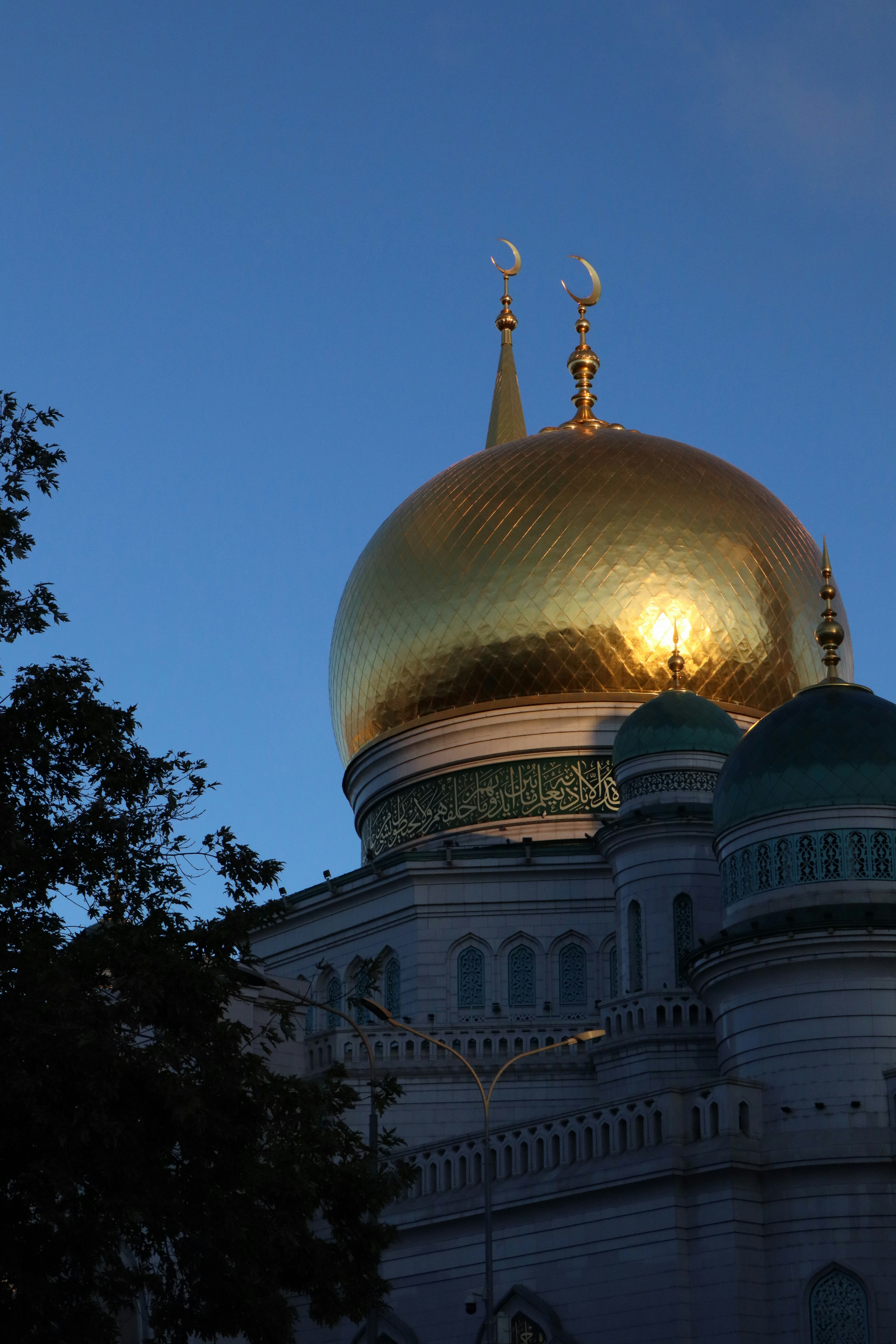 Illuminated golden domes of a mosque reflect the evening light against a deep blue sky. The intricate details of the architecture stand out amidst the surrounding foliage.
