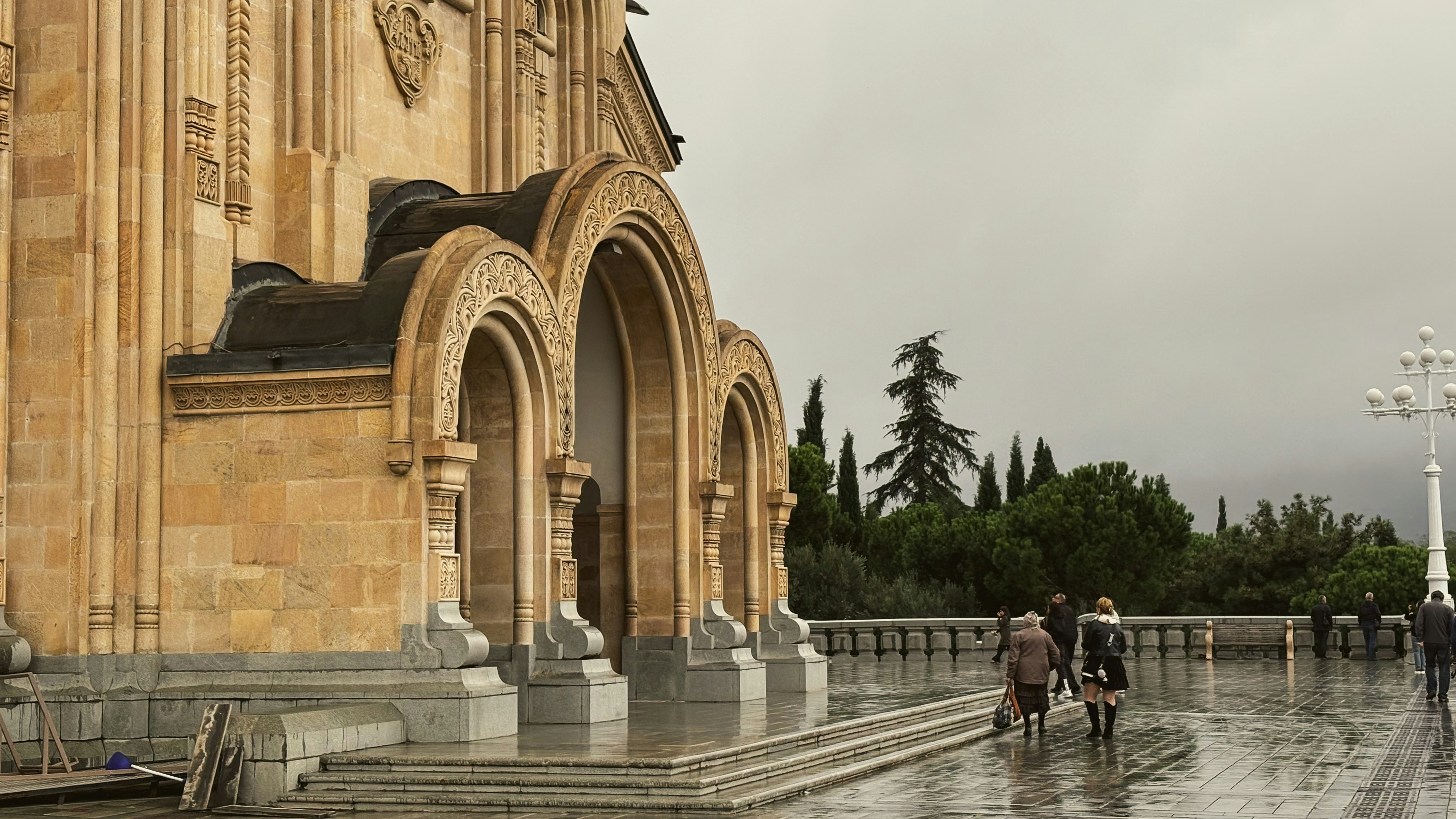 The grand arched entrance of Sameba Cathedral in Tbilisi stands over a rain-slicked plaza, where visitors walk past its ornate stonework under a moody gray sky.