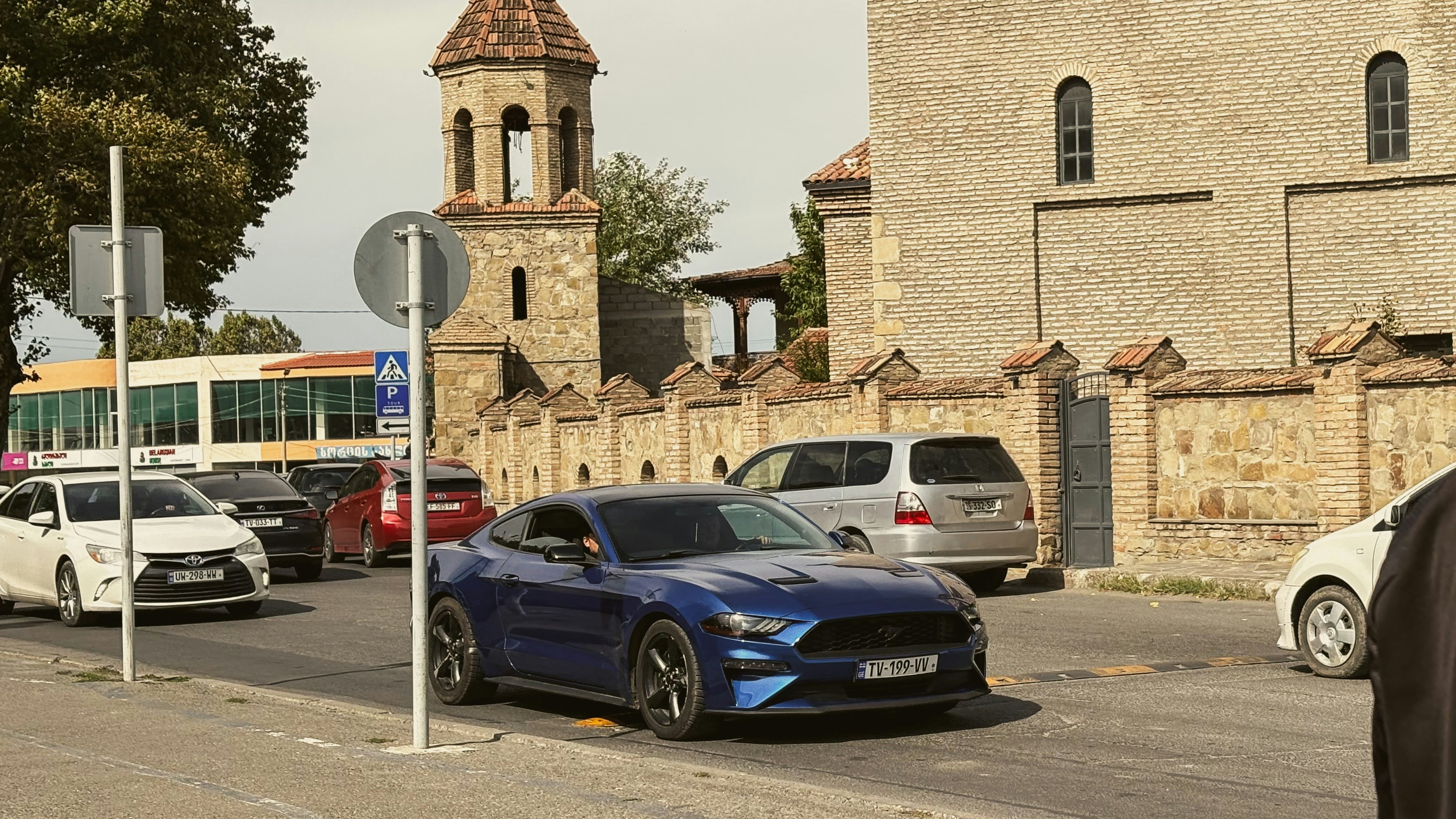 A lively street scene in Gori, Georgia, where modern cars, including a striking blue Mustang, pass by a traditional stone church and bell tower, showcasing the city’s blend of history and modern life. | Blue sports car driving on a street.