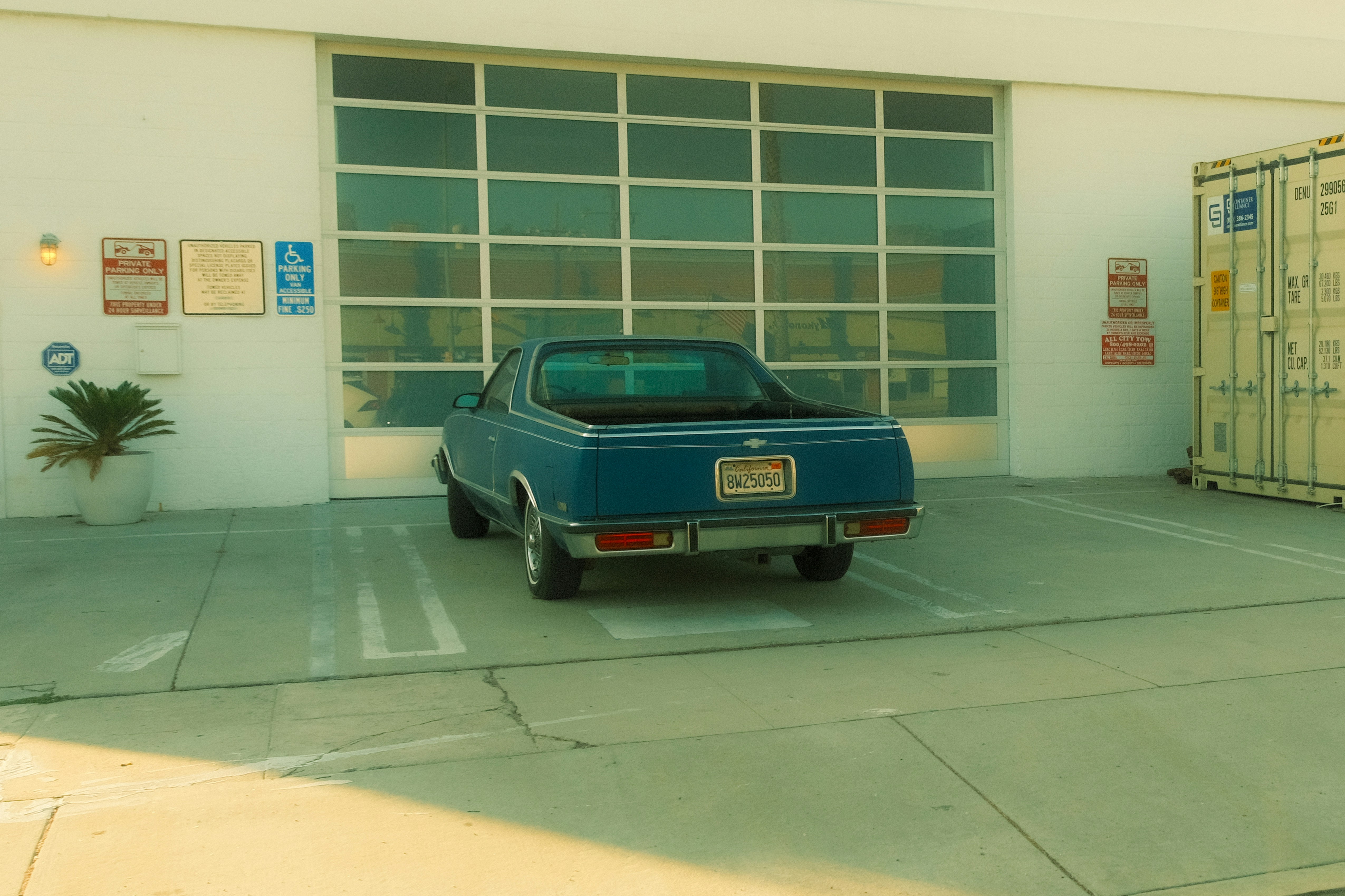 A vintage blue car parked in front of a modern garage with large glass doors, surrounded by signage and a shipping container.