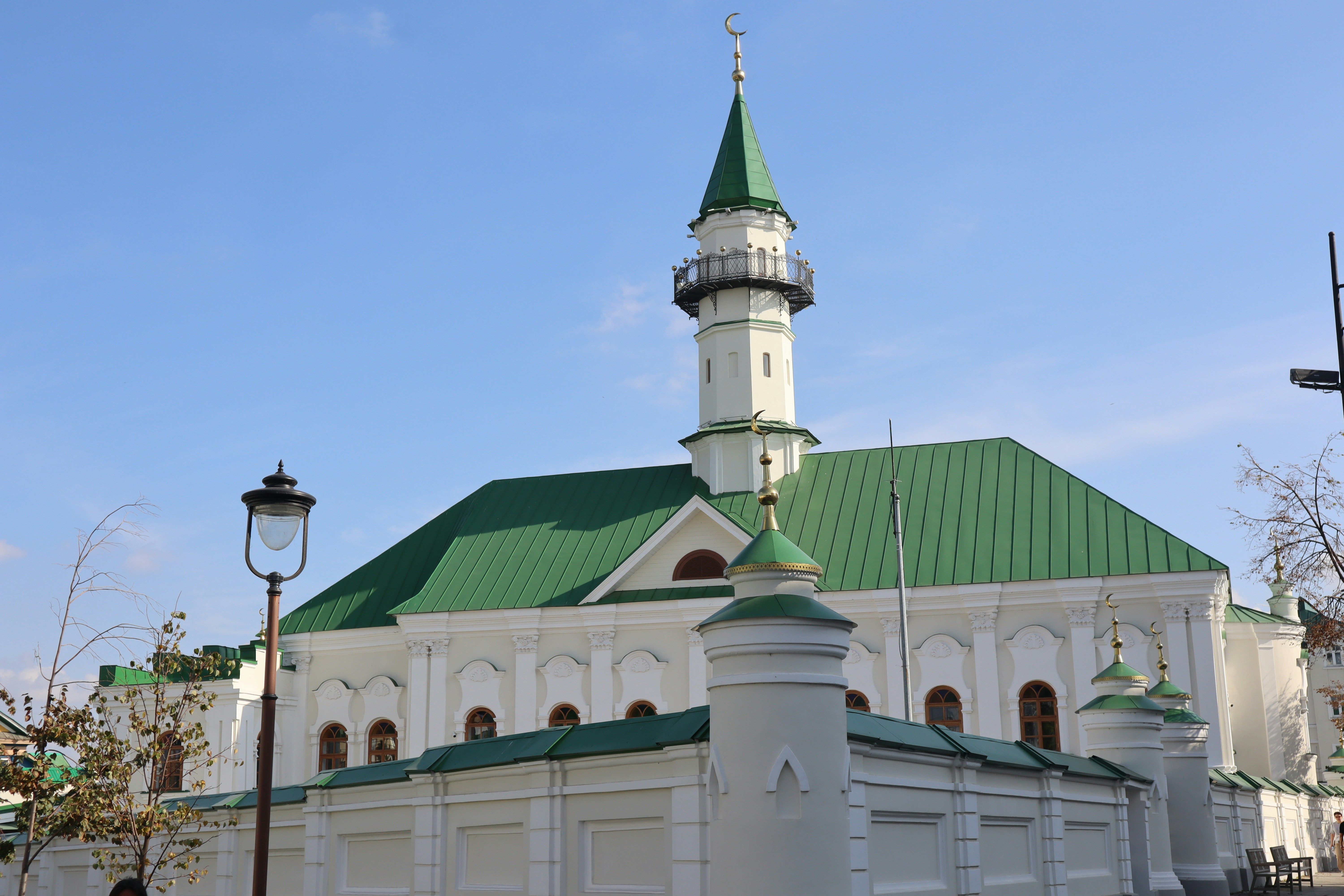 A mosque featuring a striking green roof and white walls, surrounded by urban elements. The architectural details reflect cultural significance and community presence.
