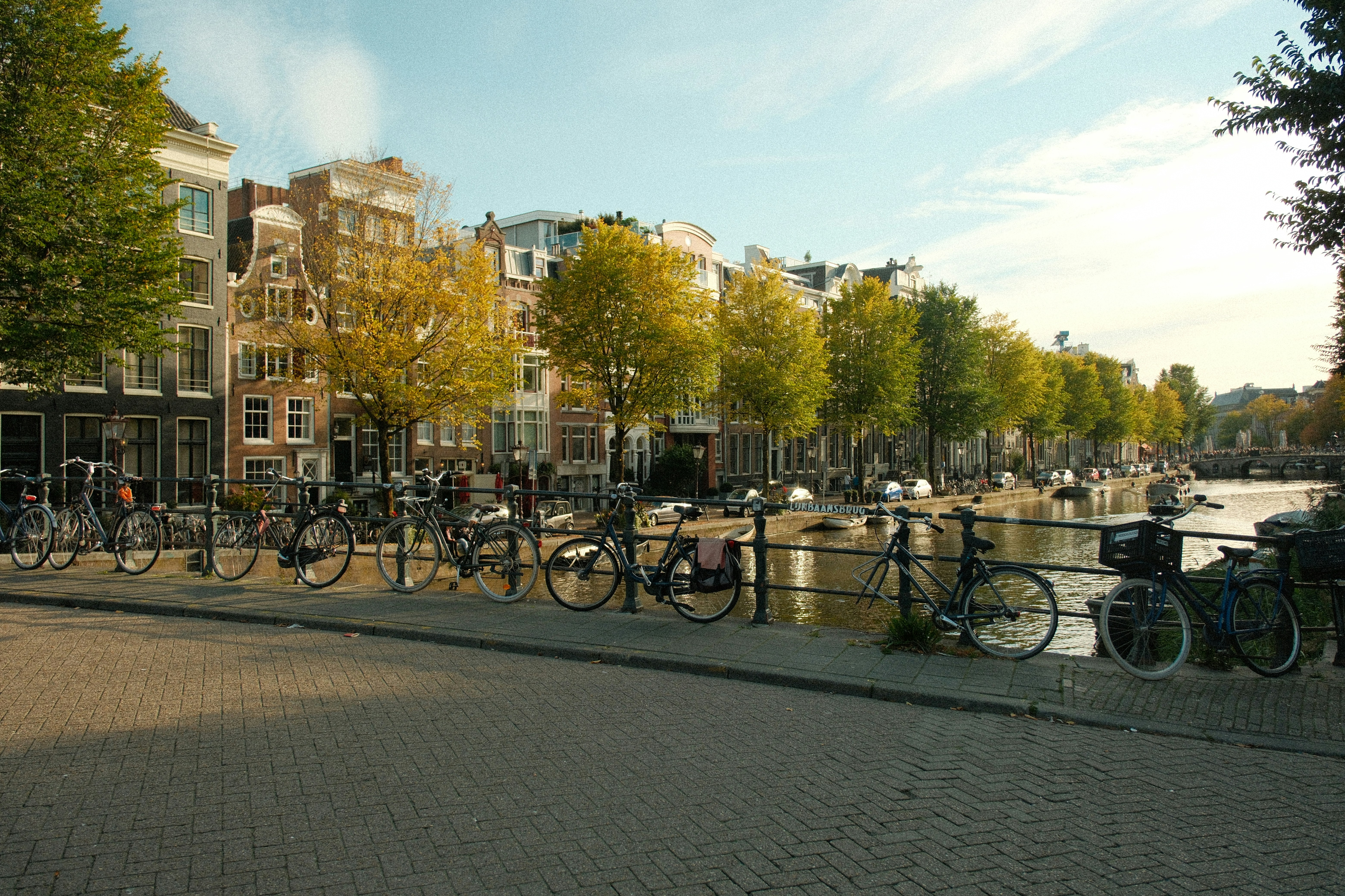 Bicycles lined along a canal in Amsterdam, framed by autumn foliage and historic architecture. The scene captures the tranquil essence of city life.